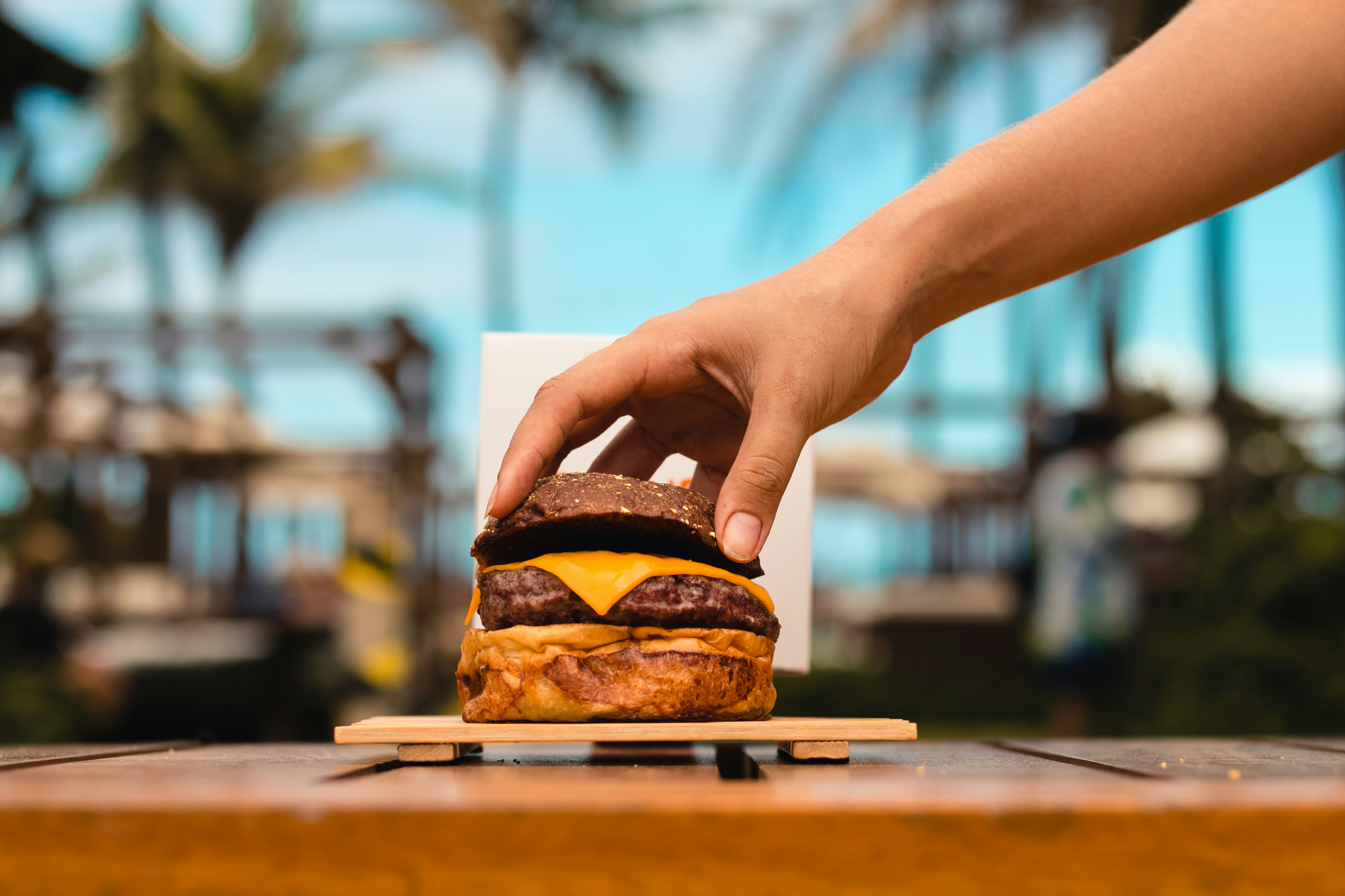 A hand reaching for a cheeseburger on a wooden table photo – Free Food ...