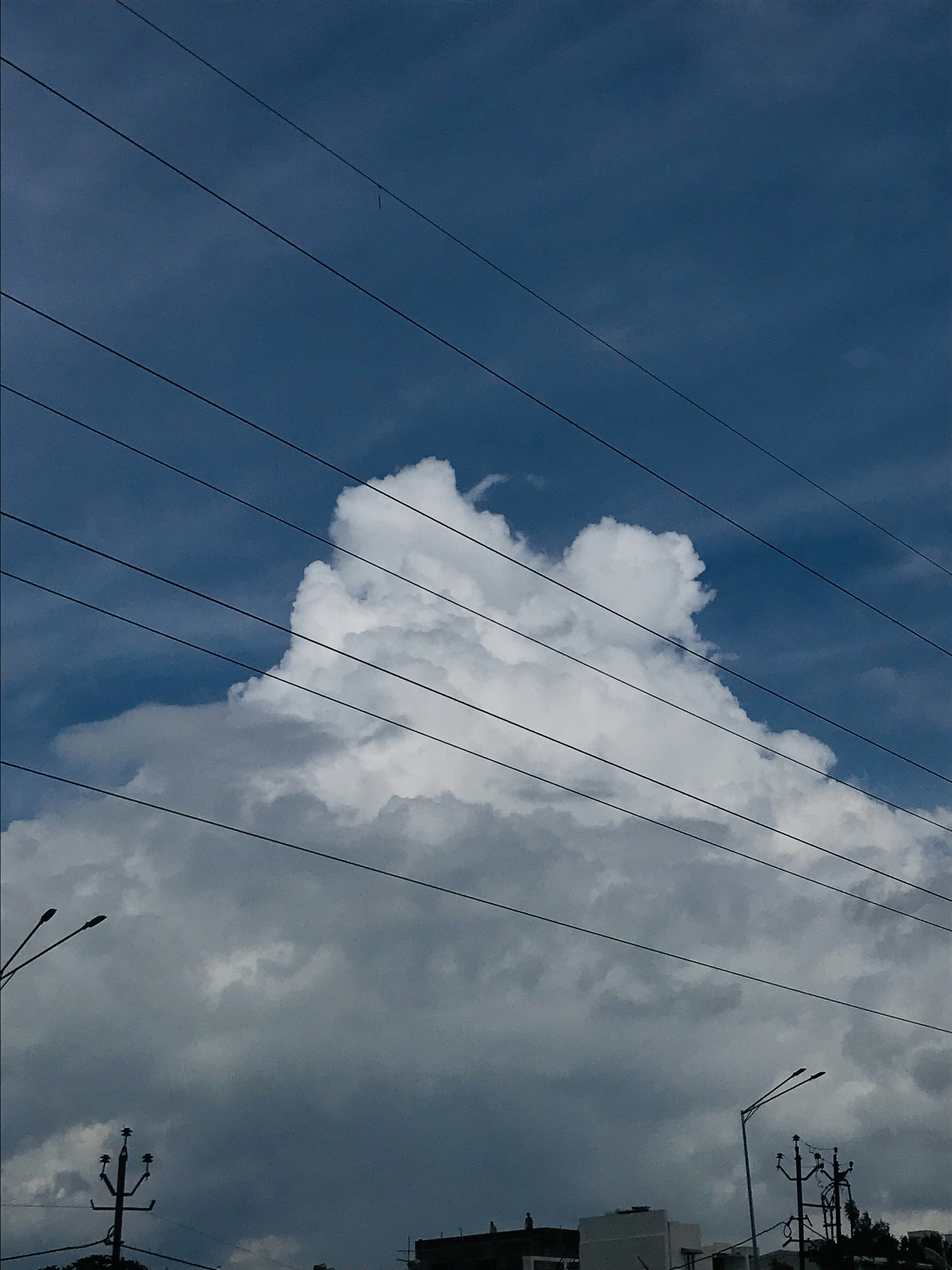 Fluffy white clouds billowing against a vibrant blue sky, intersected by power lines. The scene captures a moment of serene weather.
