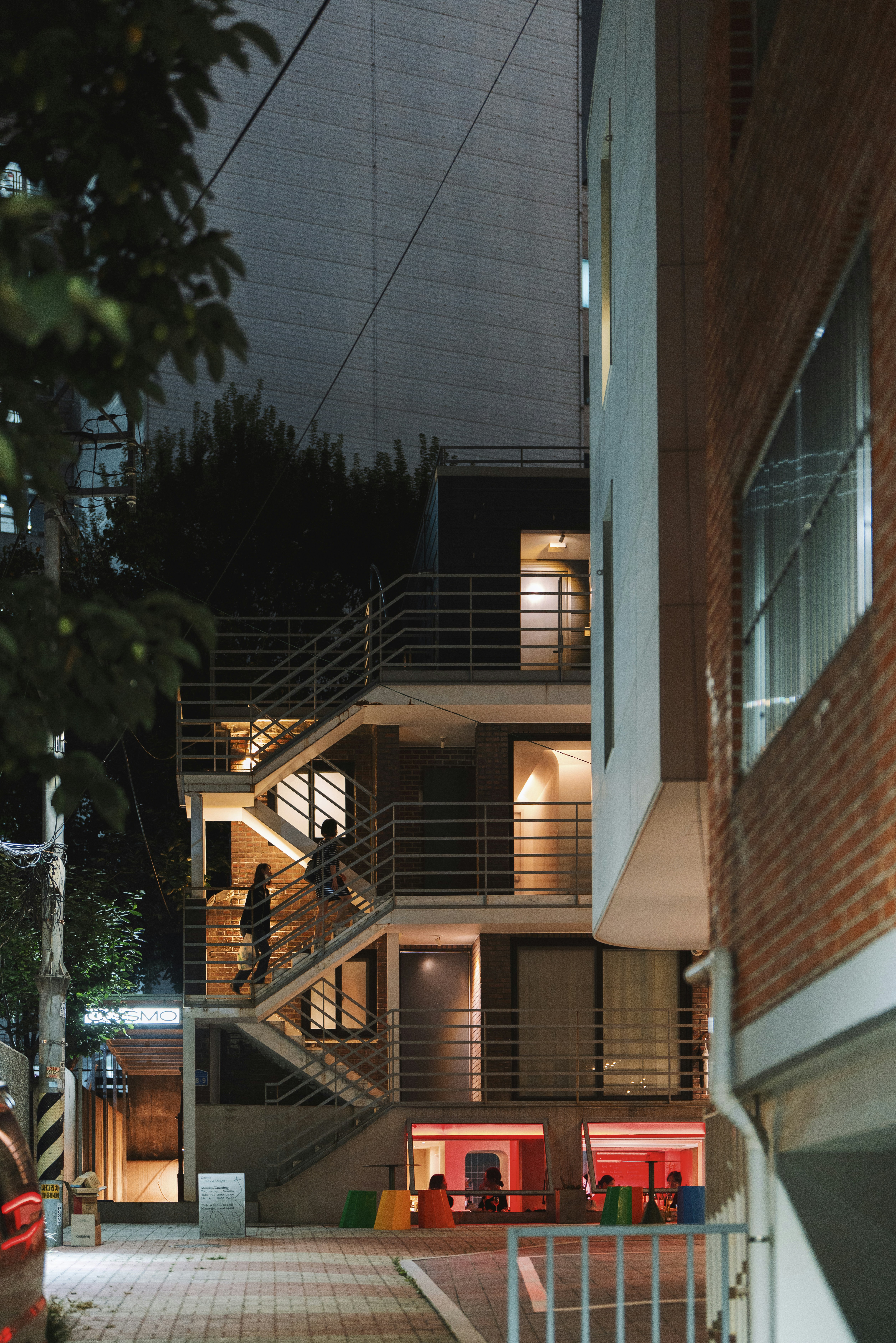 A person ascends a staircase of a modern building illuminated by warm lights, surrounded by lush greenery and urban elements.