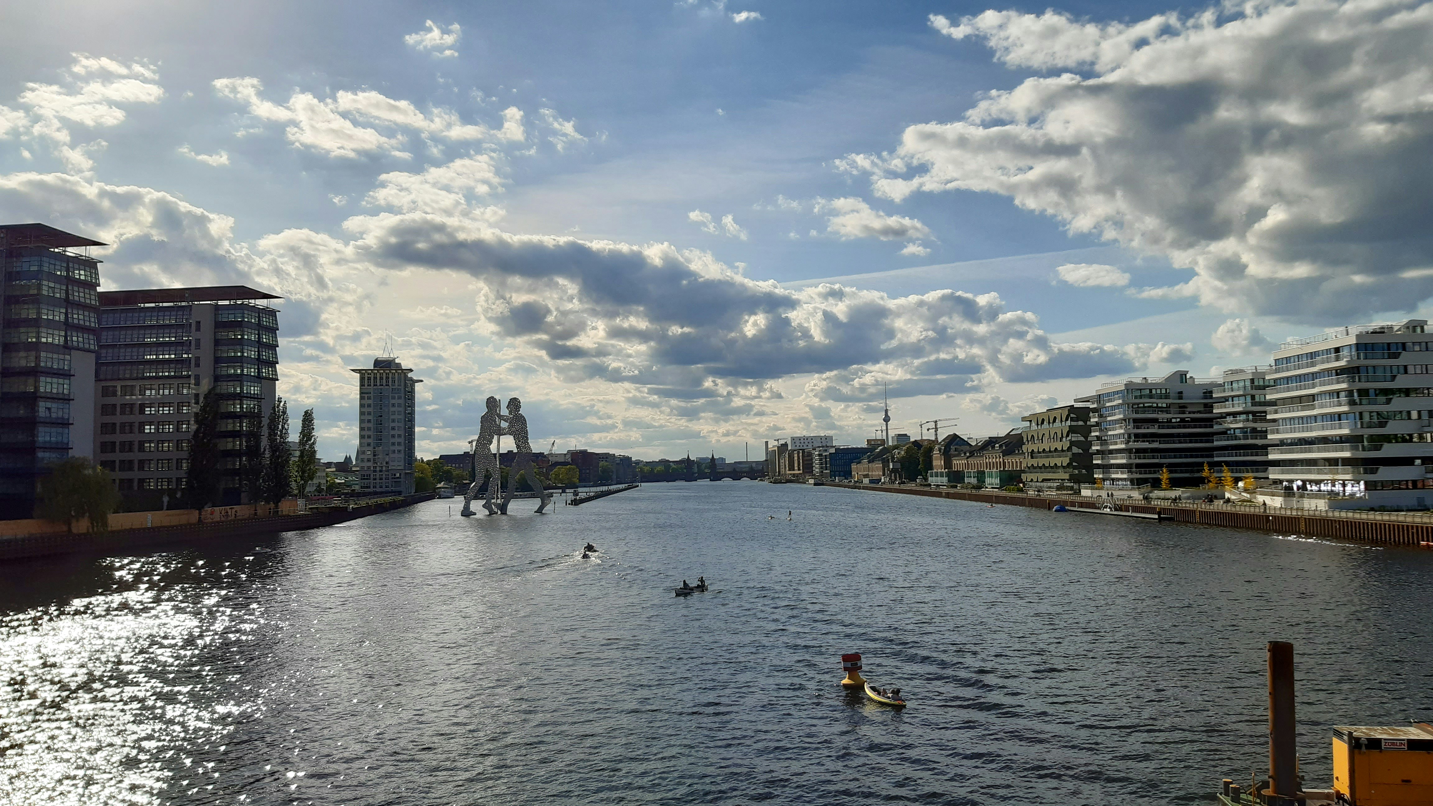 A serene riverscape featuring modern architecture alongside a sculpture, with boats gliding through the water under a partly cloudy sky.