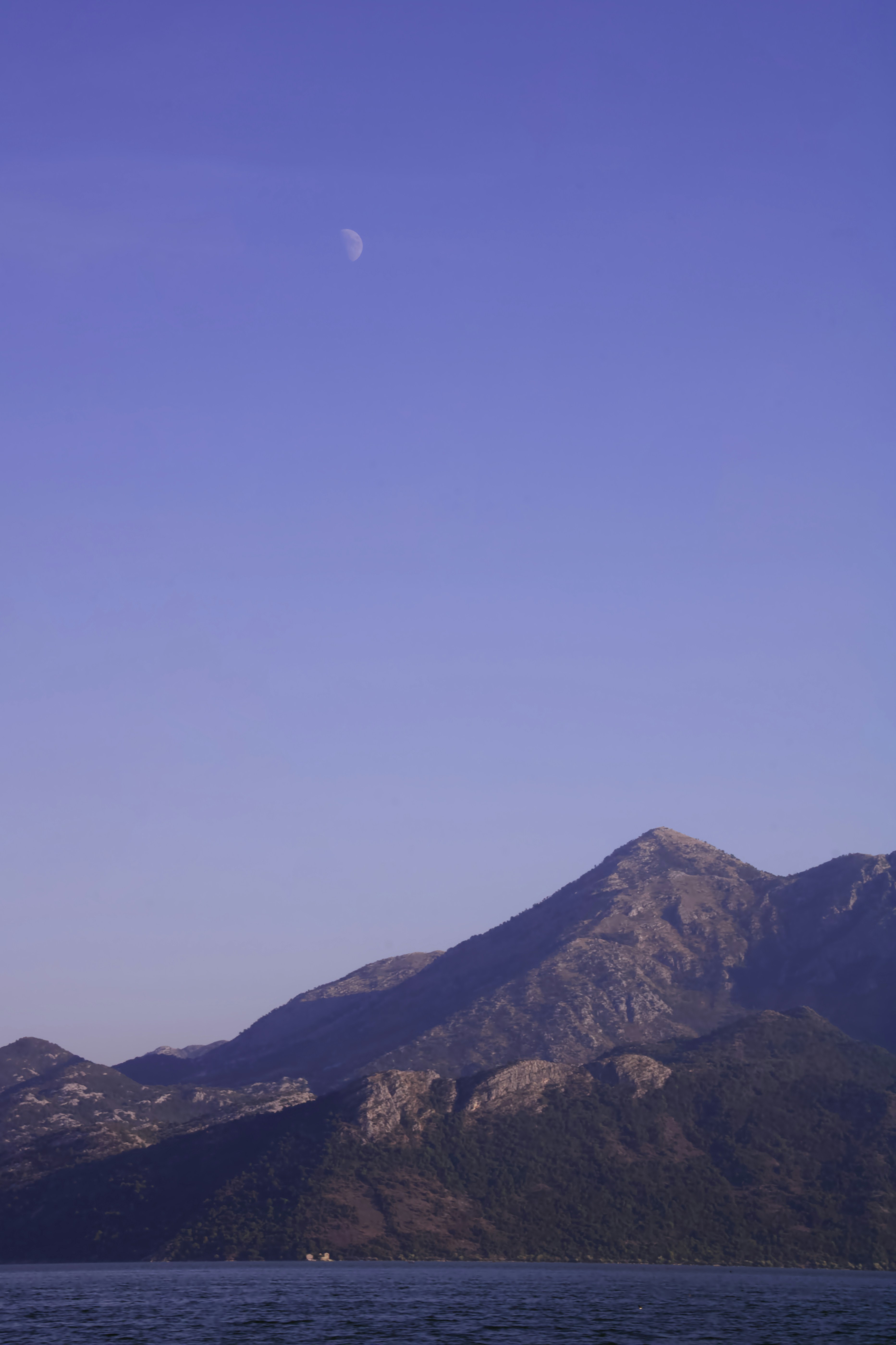 A serene mountain landscape under a clear sky, with a crescent moon visible above the peaks. The tranquil waters reflect the natural beauty of the scene.