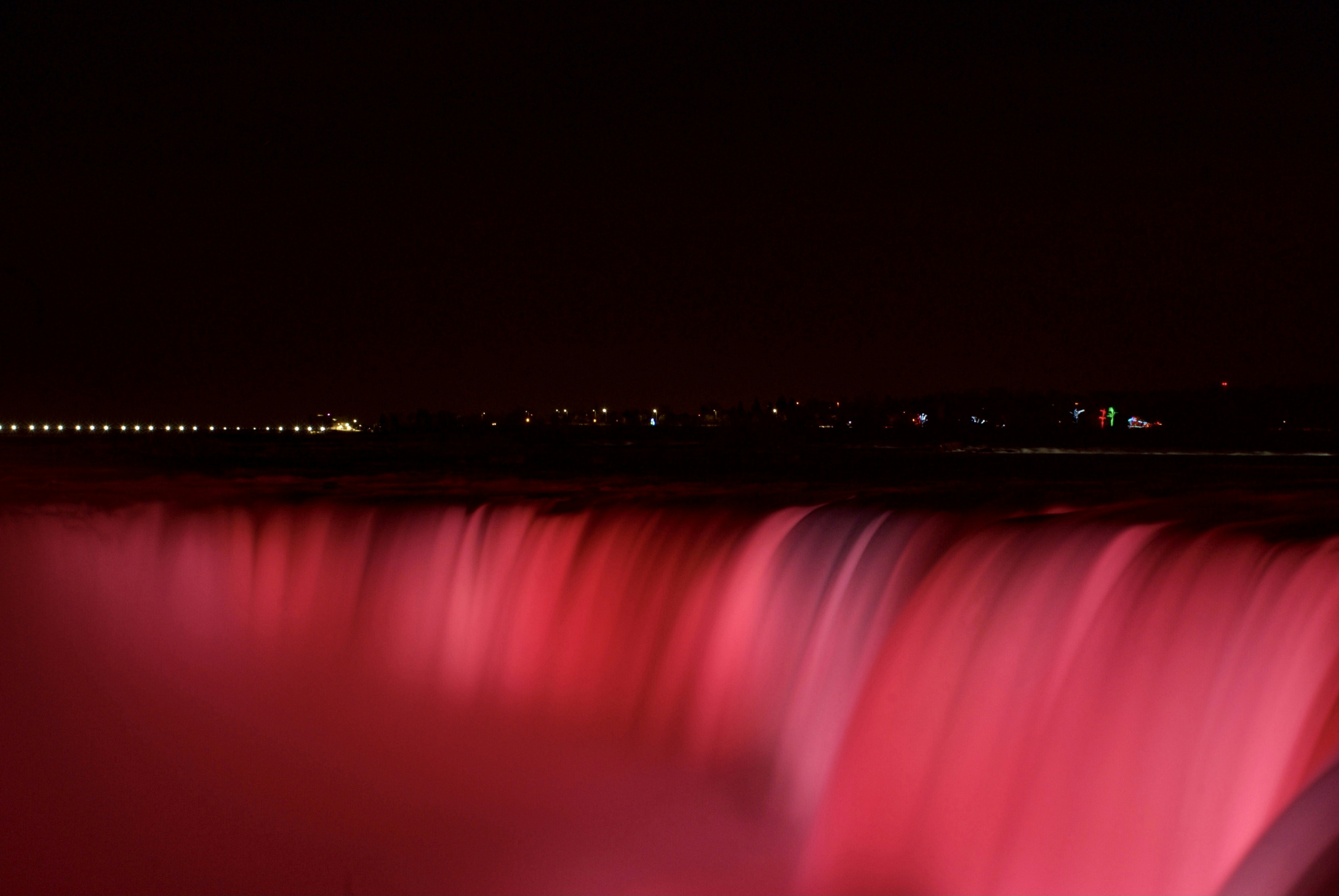 Long-exposure night photograph of a neon-lit waterfall bathed in red hues, with a distant skyline glow.