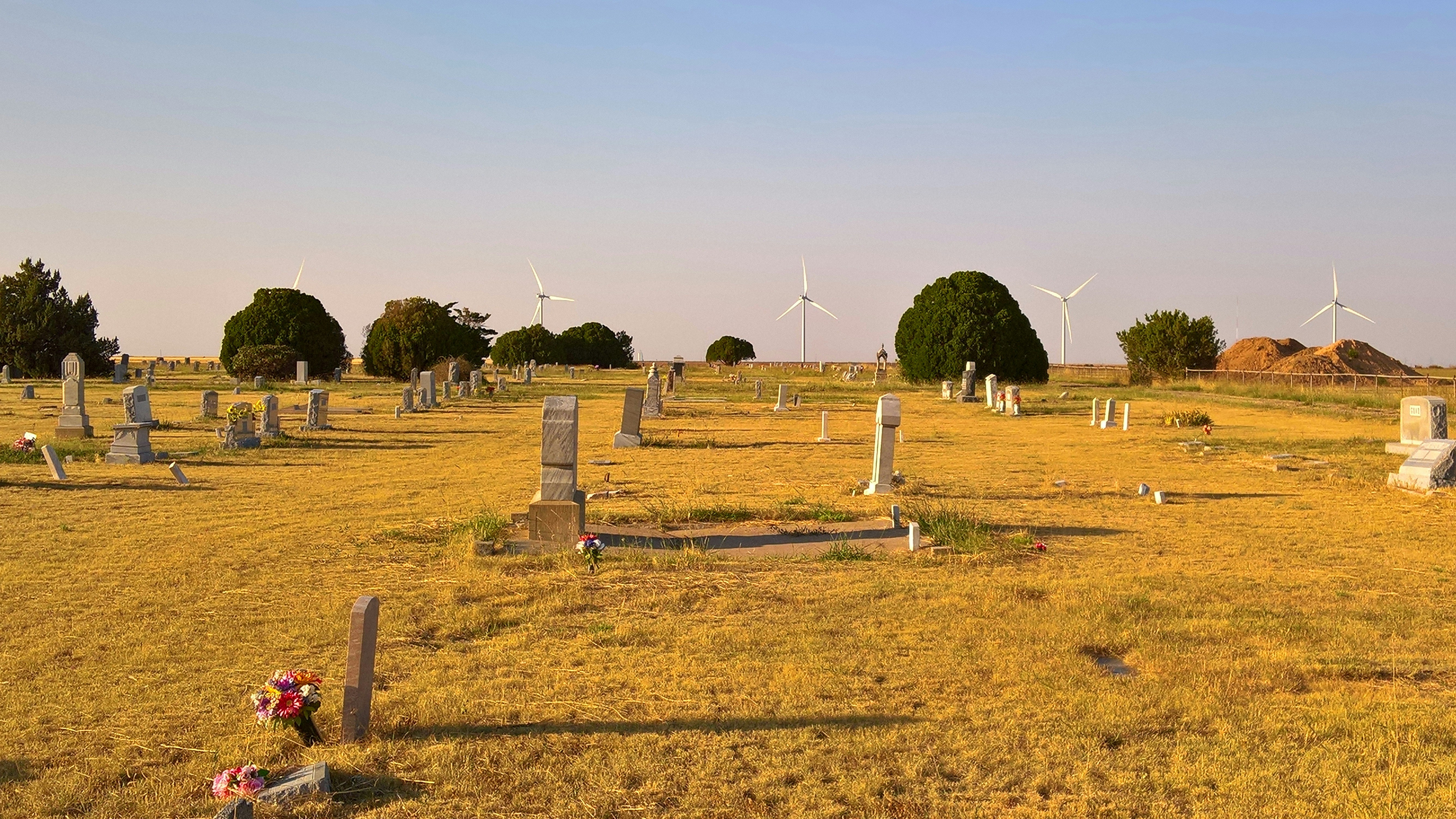 A serene cemetery landscape dotted with gravestones, surrounded by wind turbines under a clear sky. Flowers adorn some graves, adding a touch of color to the scene.