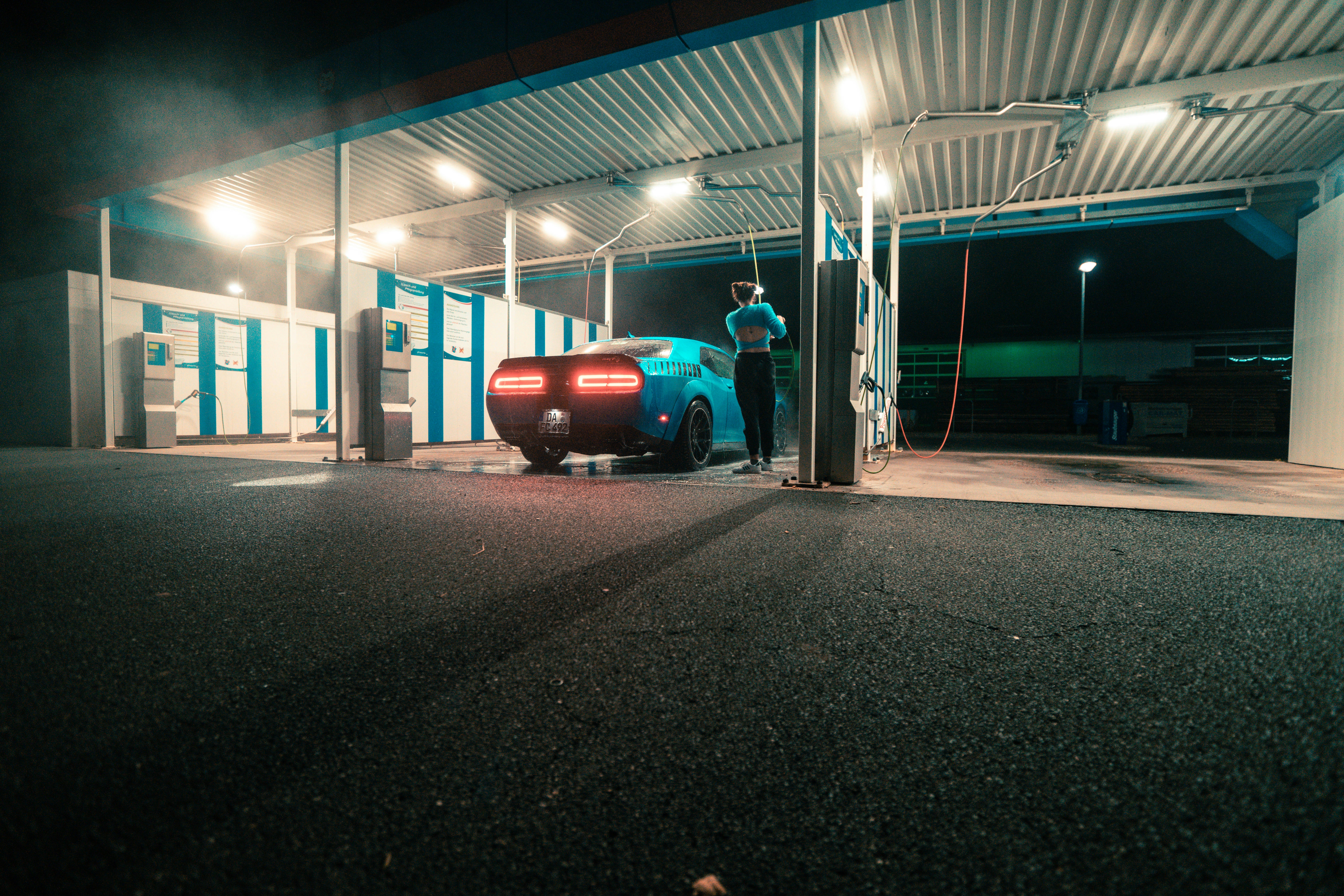 Person washing a sports car under bright lights at an empty car wash at night.