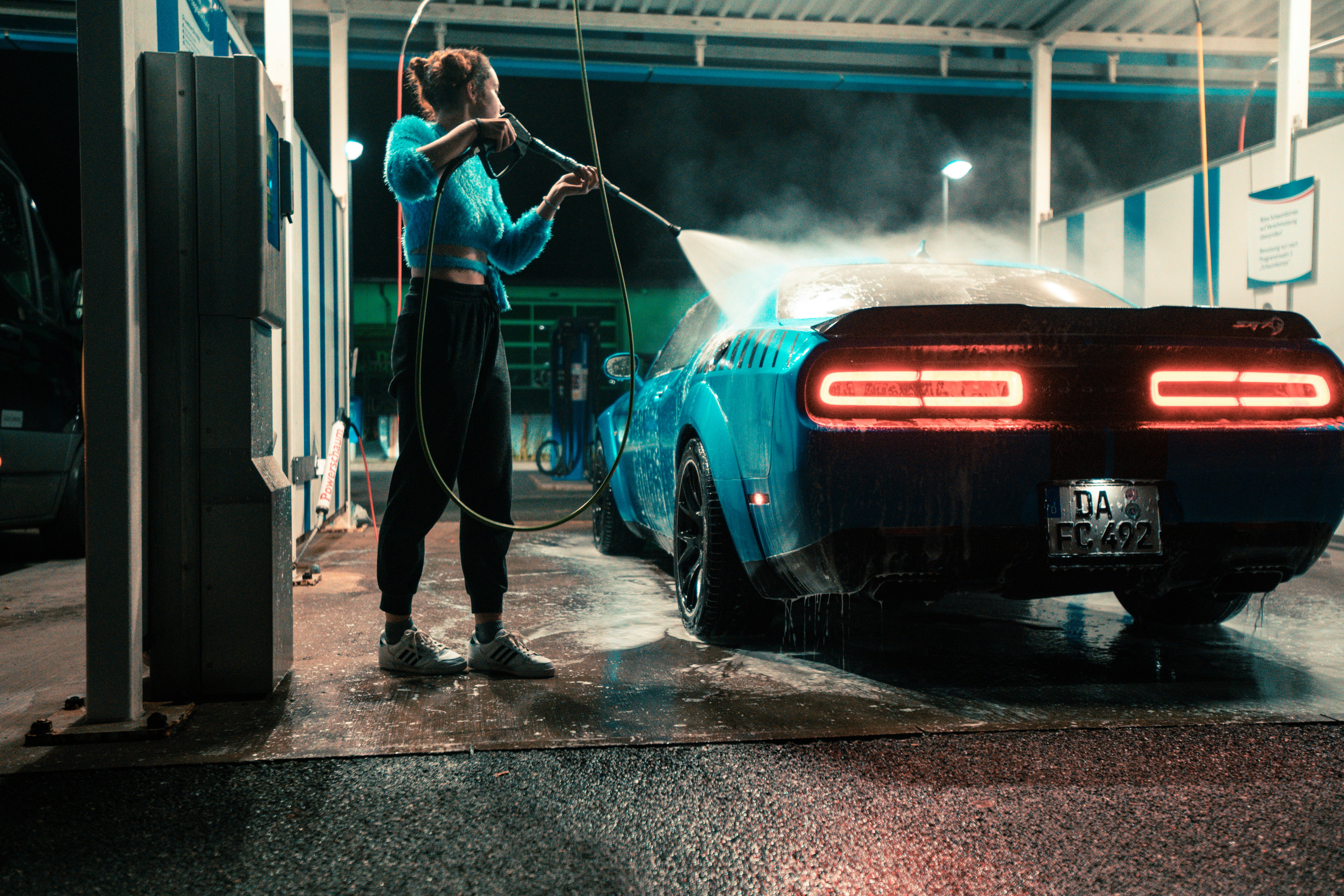 A person washes a blue sports car at a nighttime car wash, illuminated by bright lights and surrounded by mist from the water spray.
