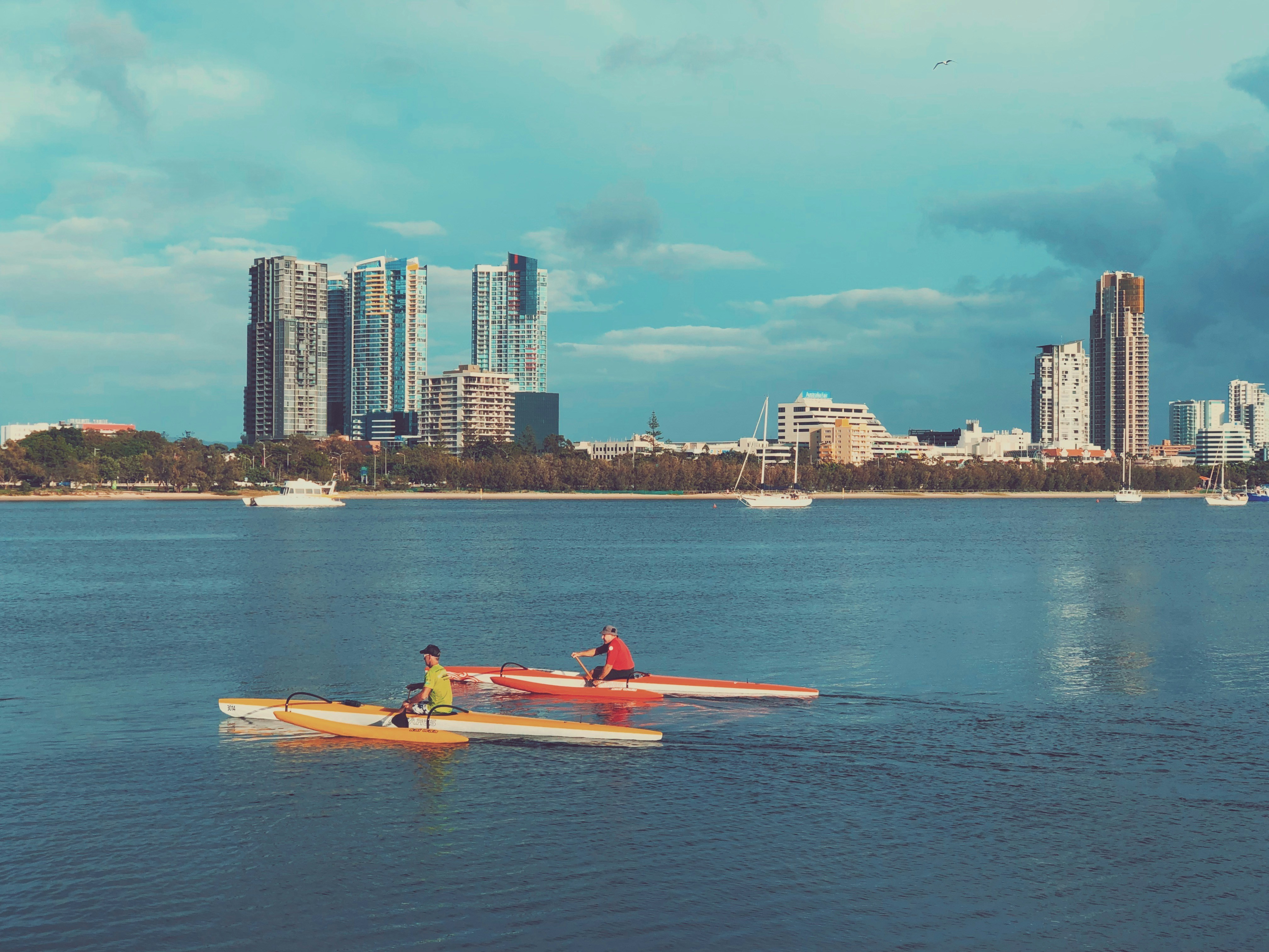 Two kayakers glide across a calm river, framed by a backdrop of modern skyscrapers and a clear blue sky.