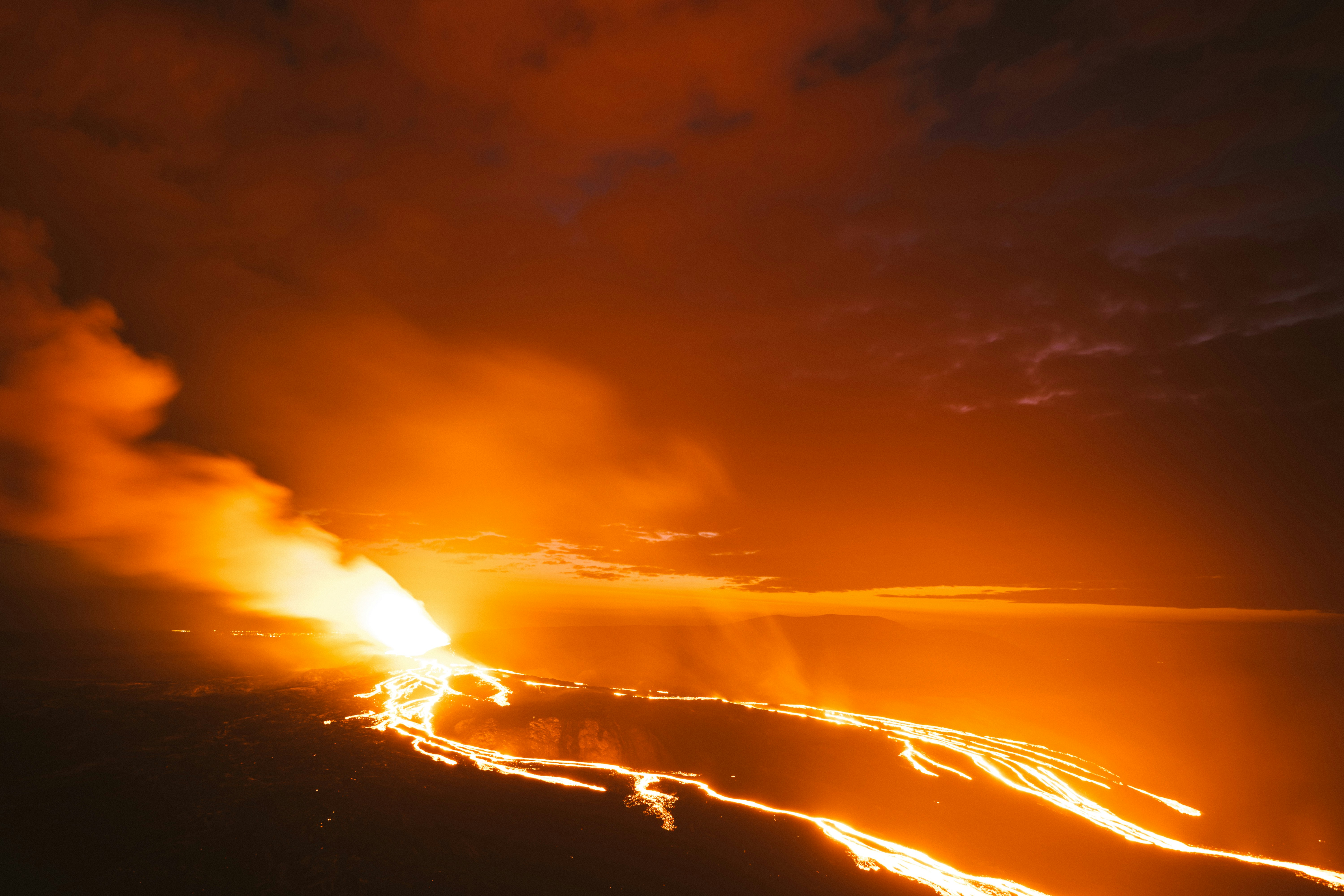Volcanic eruption with flowing lava illuminating the dark landscape and clouds above. The fiery glow contrasts against the night sky.