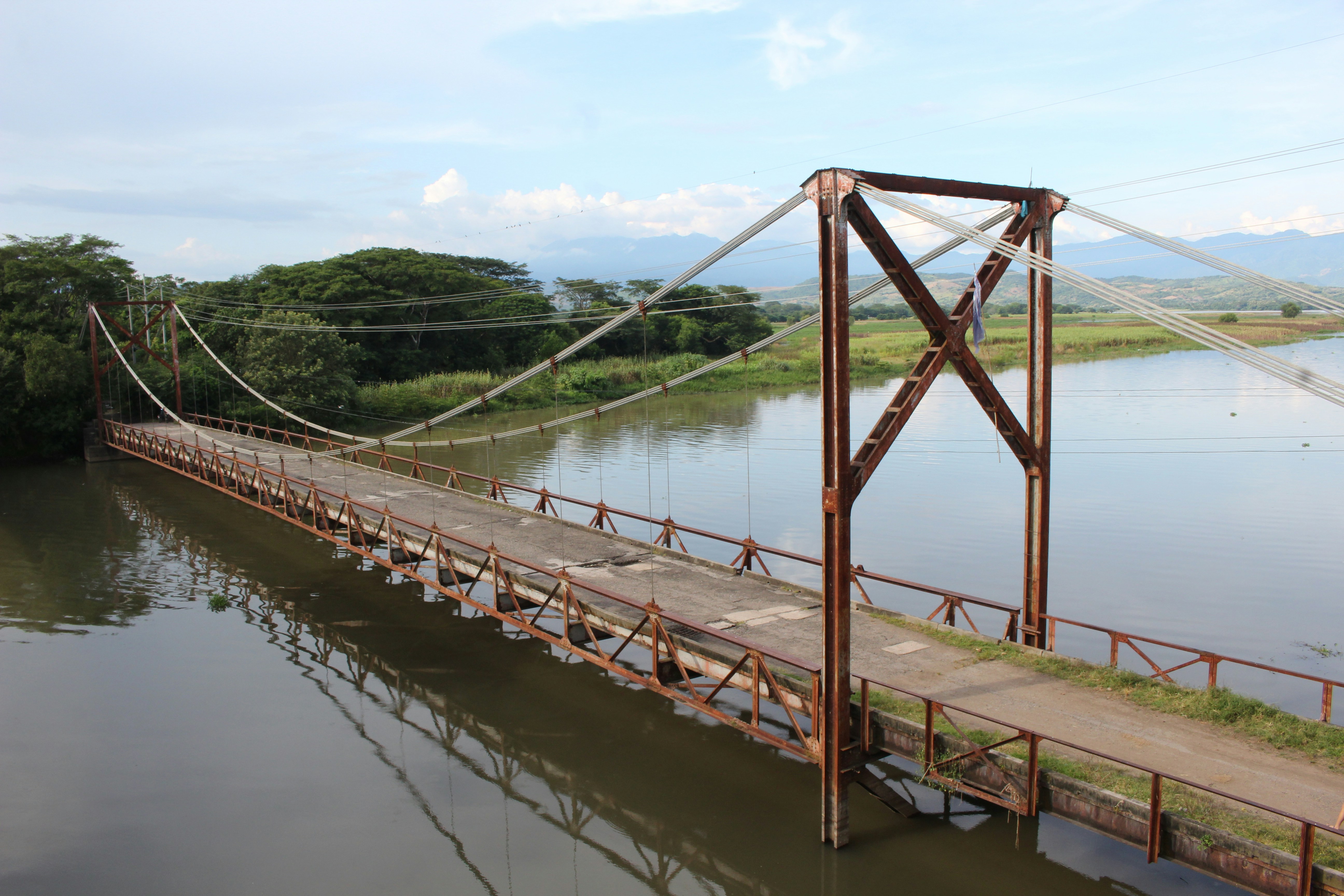A suspension bridge spans a calm river, framed by lush greenery and distant mountains under a clear sky.