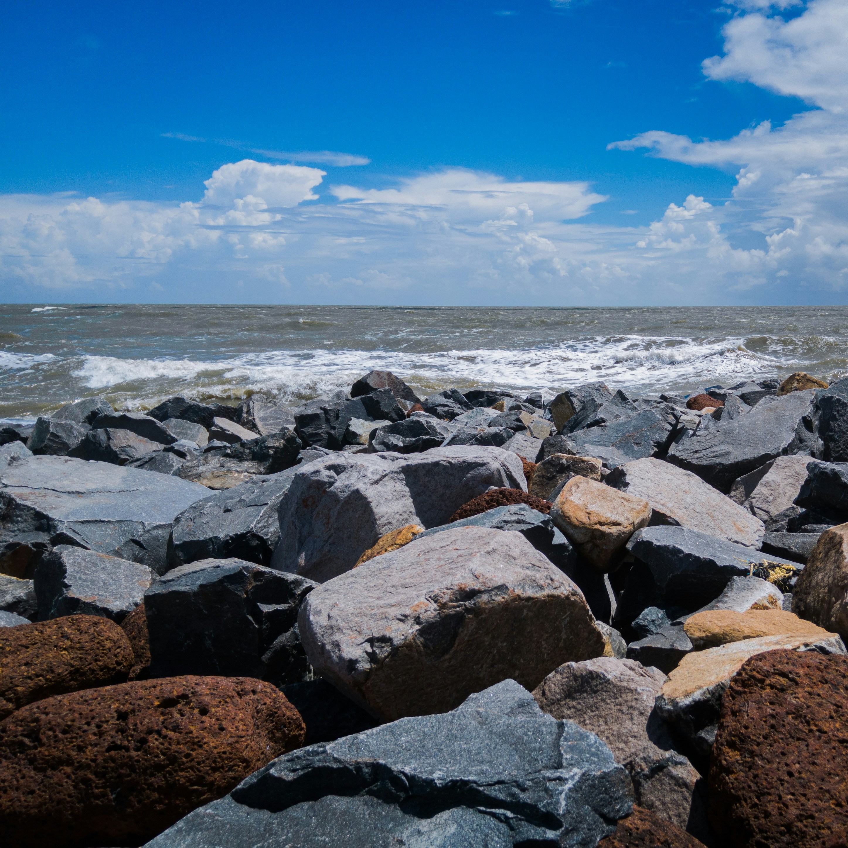 Waves crash against a rugged shoreline of assorted rocks under a bright blue sky filled with fluffy clouds.