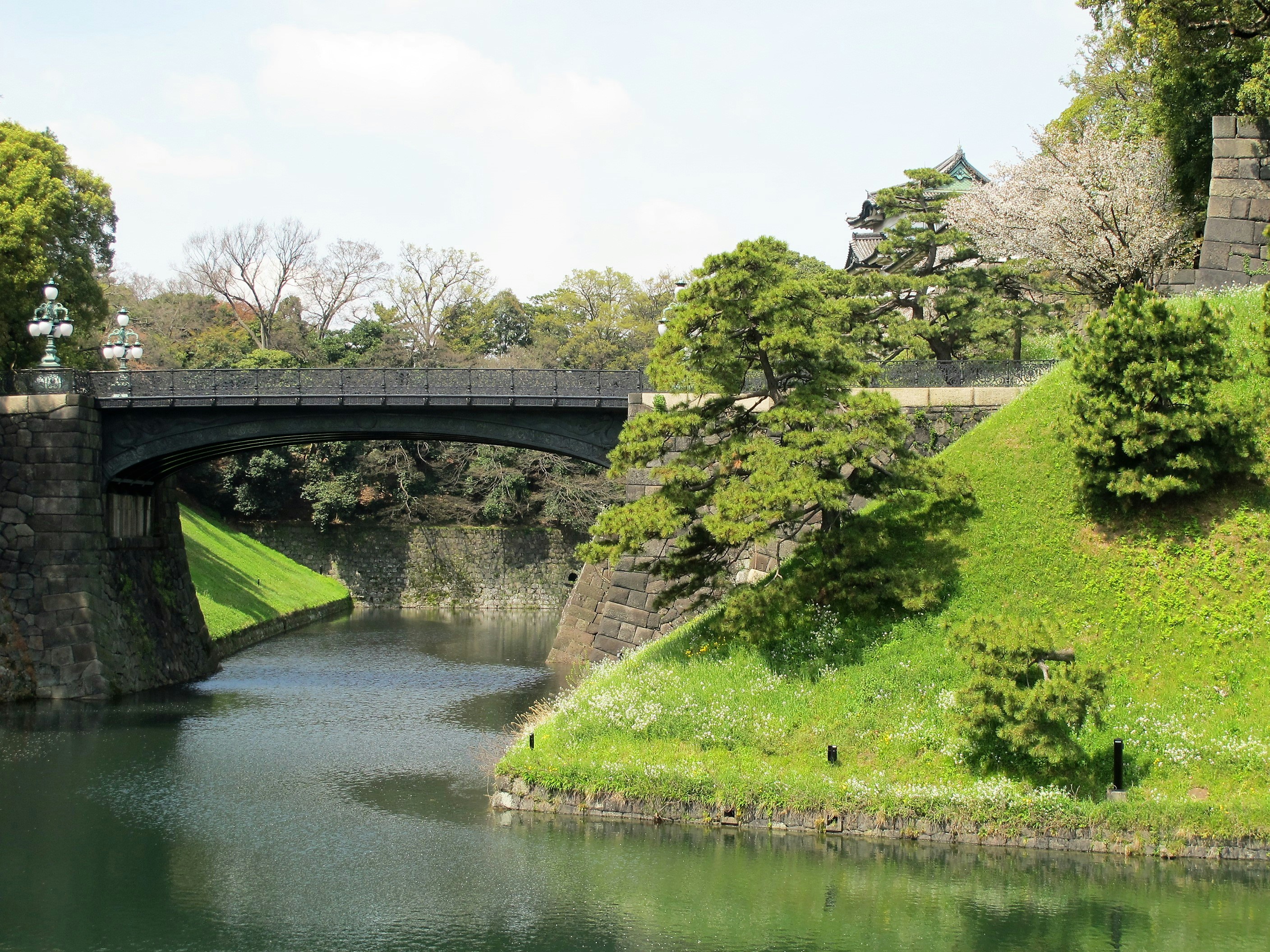 A historic stone bridge arches over a tranquil moat, surrounded by lush greenery and blooming trees, with a glimpse of a castle in the background.