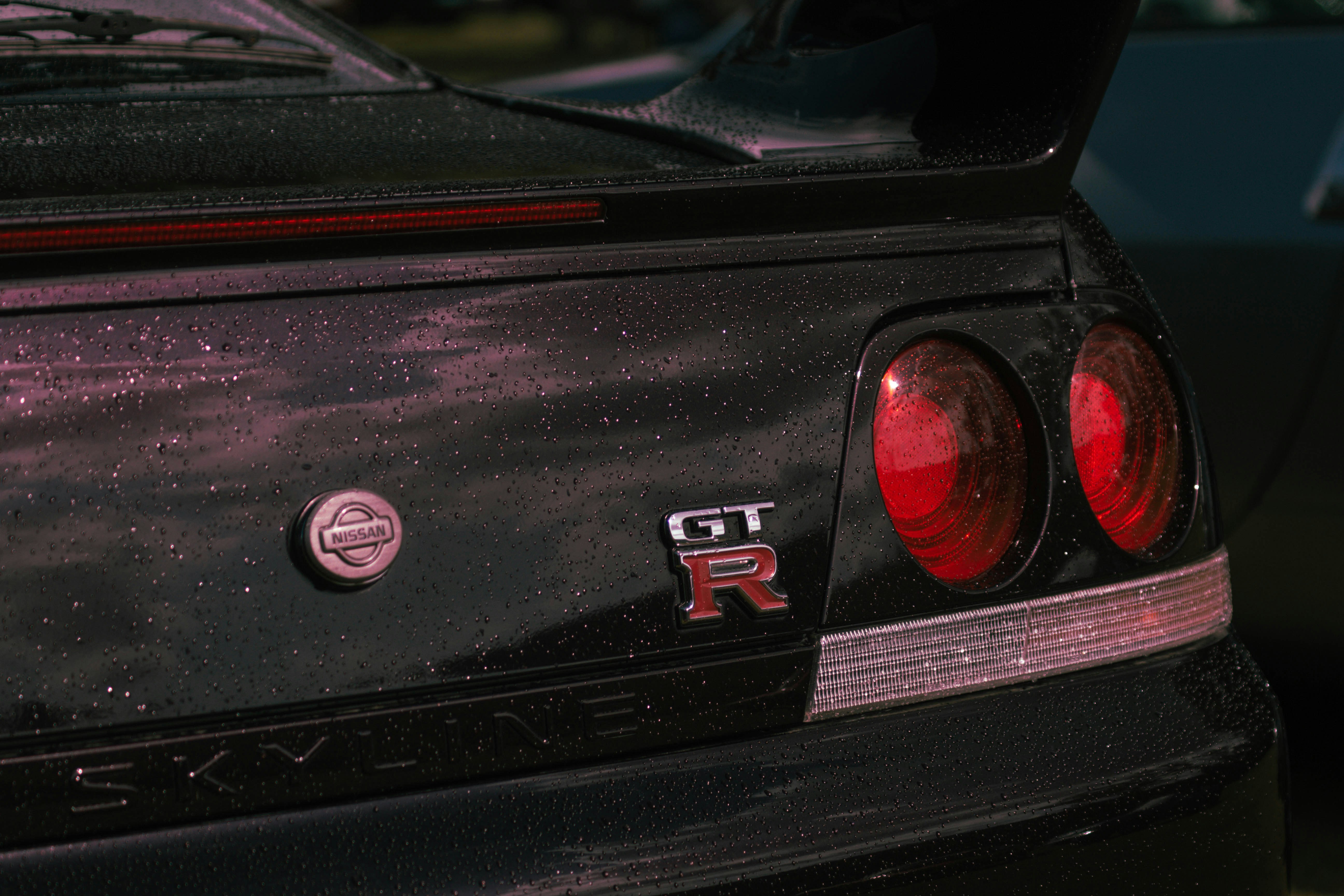 Close-up of a Nissan GT-R emblem on a sleek black car, showcasing raindrops glistening on the surface. The iconic design highlights the car's performance lineage.