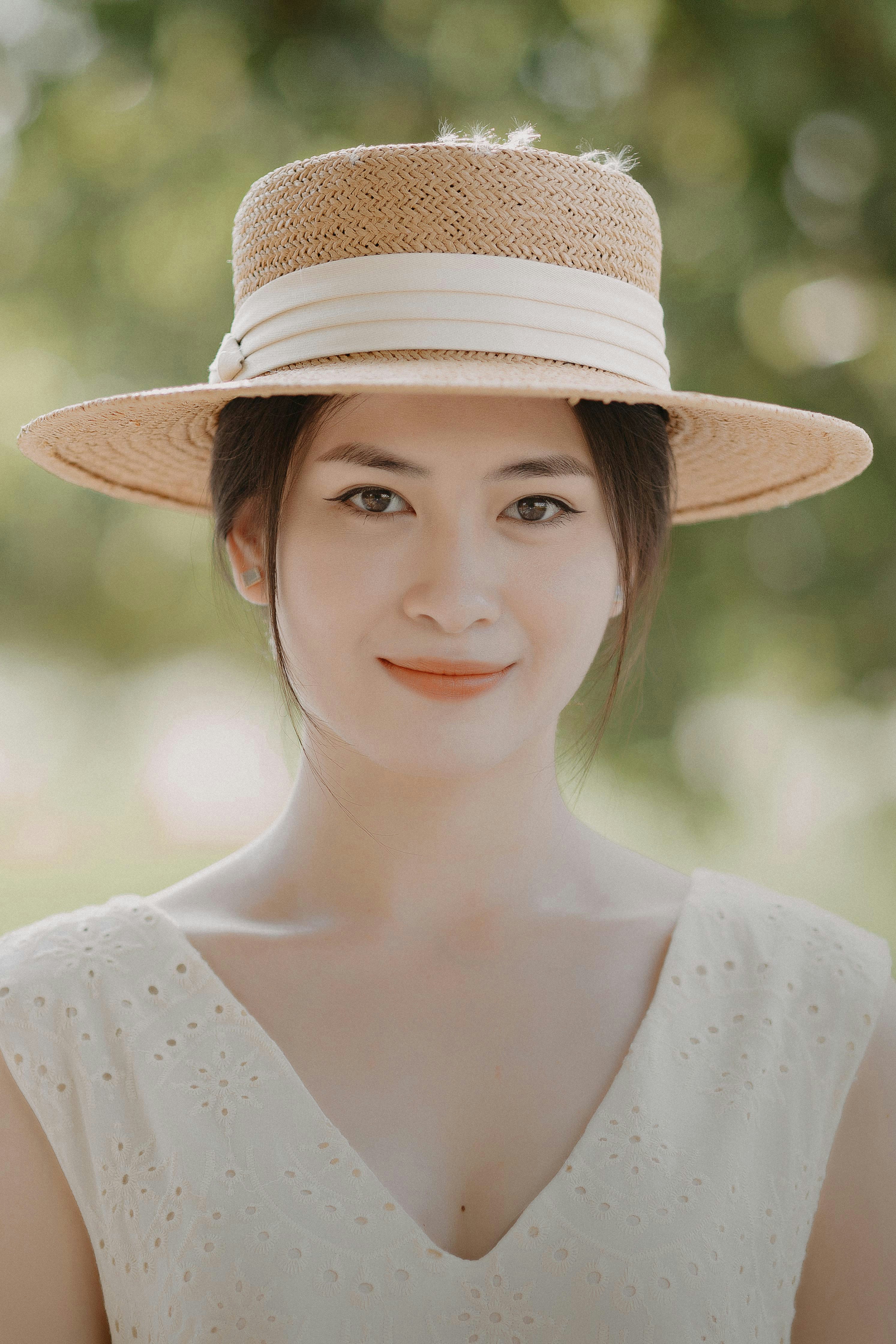 A young woman in a straw hat and white dress poses gracefully against a softly blurred background of greenery.