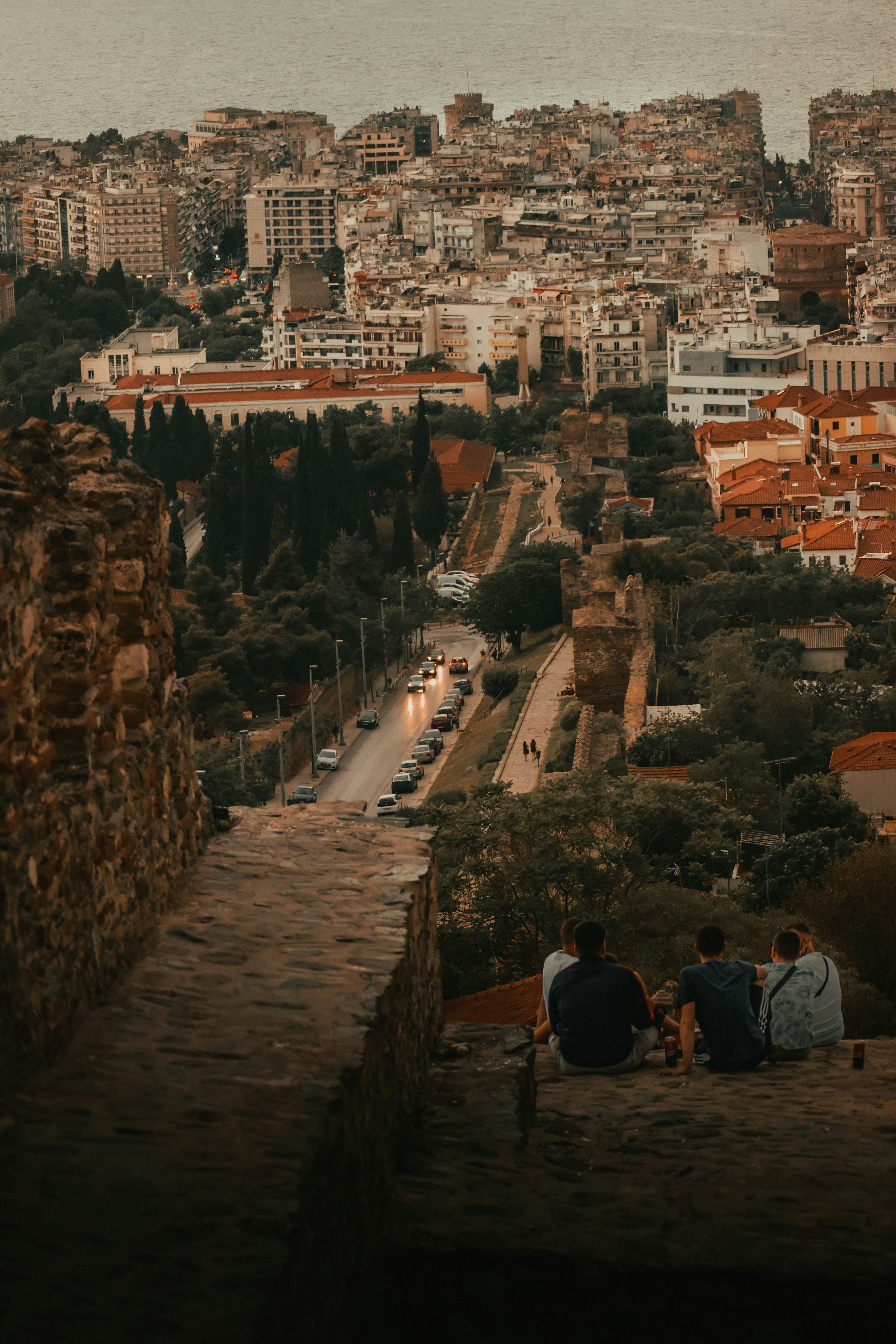 Three friends sitting on a stone ledge overlooking a sprawling cityscape at dusk, framed by ancient ruins. The scene captures a blend of nature and urban life.