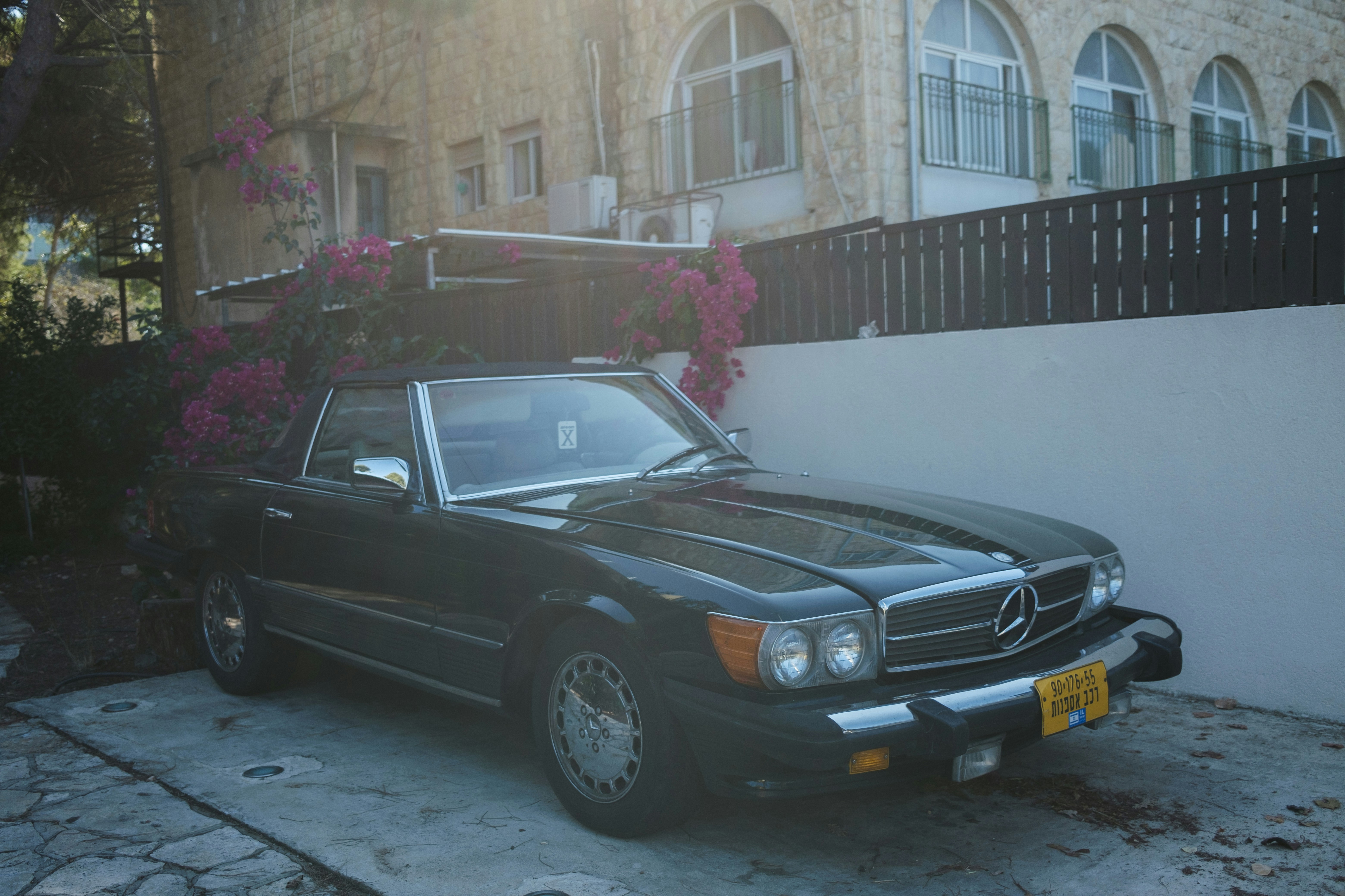 Black Mercedes-Benz W126 on a sunlit driveway, framed by pink bougainvillea and a weathered wall