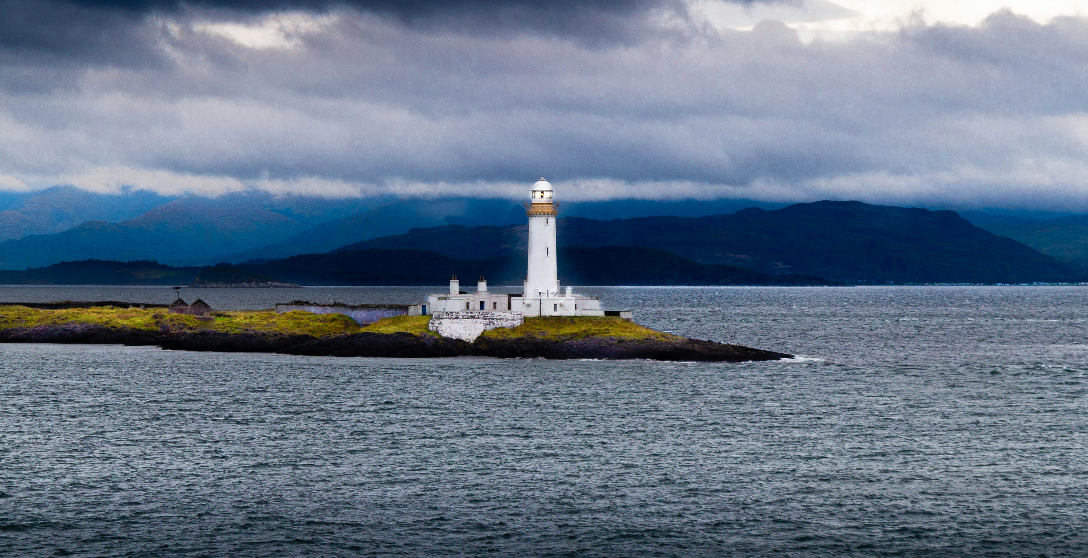 Lismore Lighthouse stands on a small island against a backdrop of mountains and a cloudy sky.