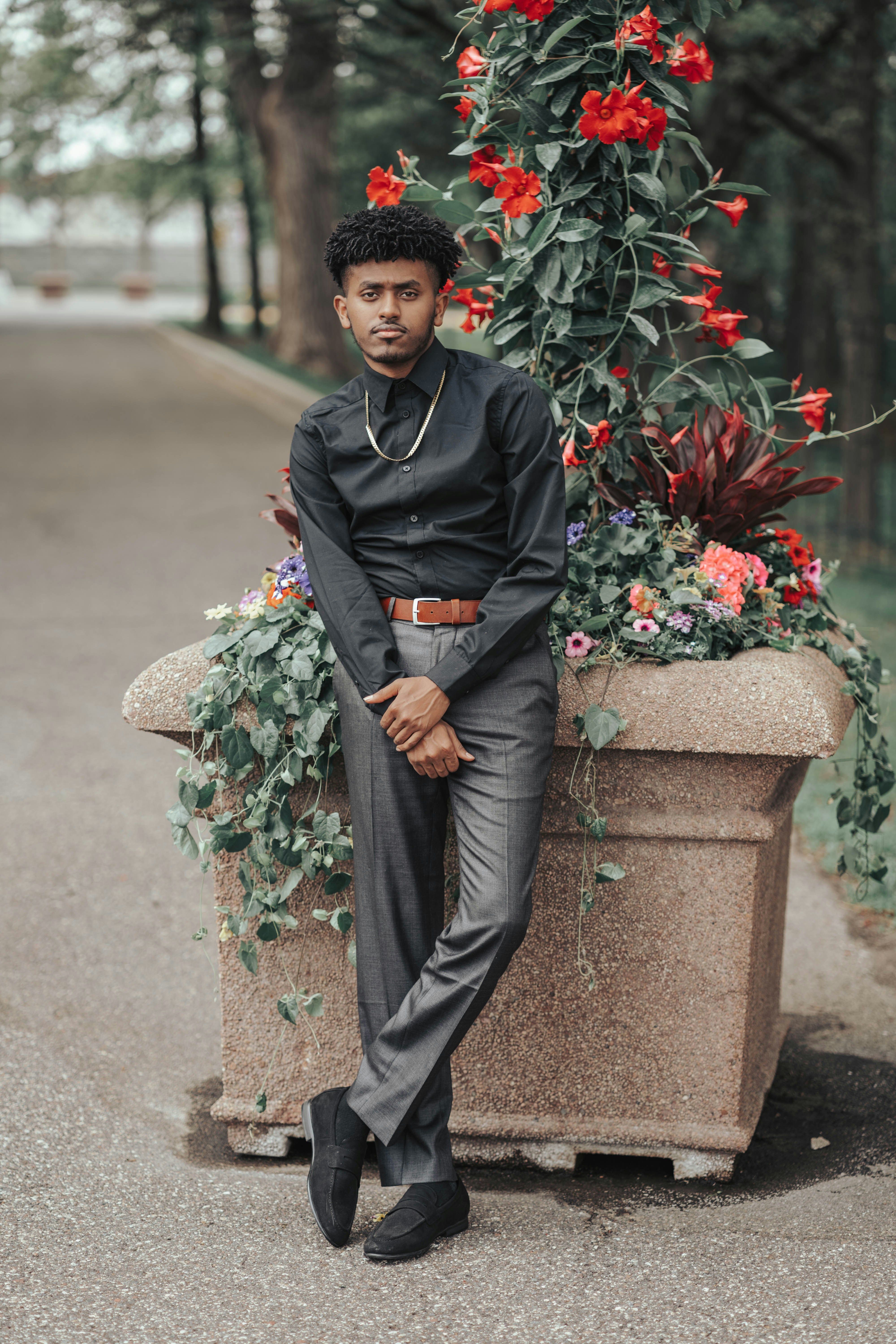 A stylish individual poses confidently beside a vibrant floral arrangement in a serene outdoor setting.