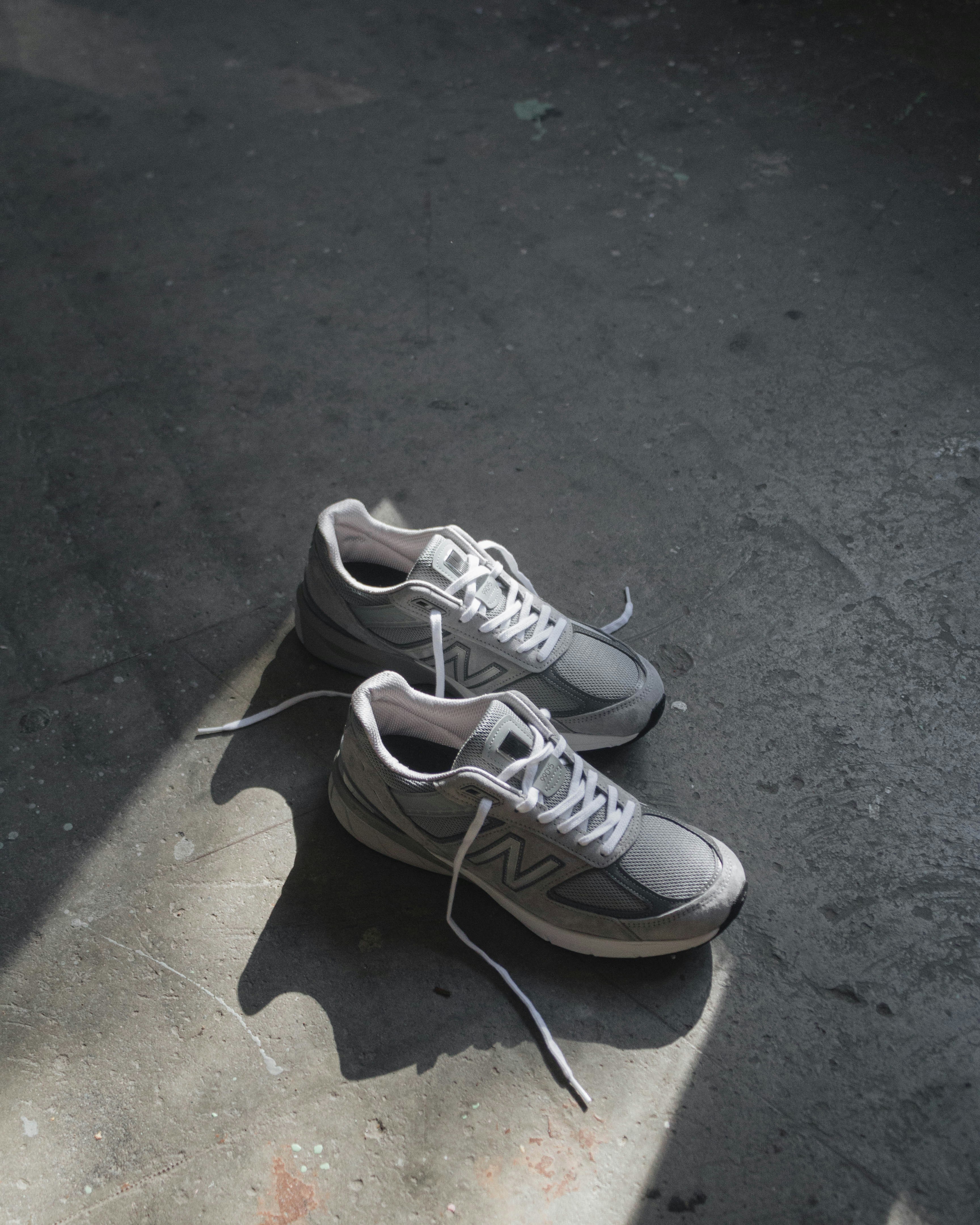A pair of gray New Balance sneakers resting on a textured concrete floor, partially illuminated by natural light. The interplay of light and shadow highlights the shoes' design and details.