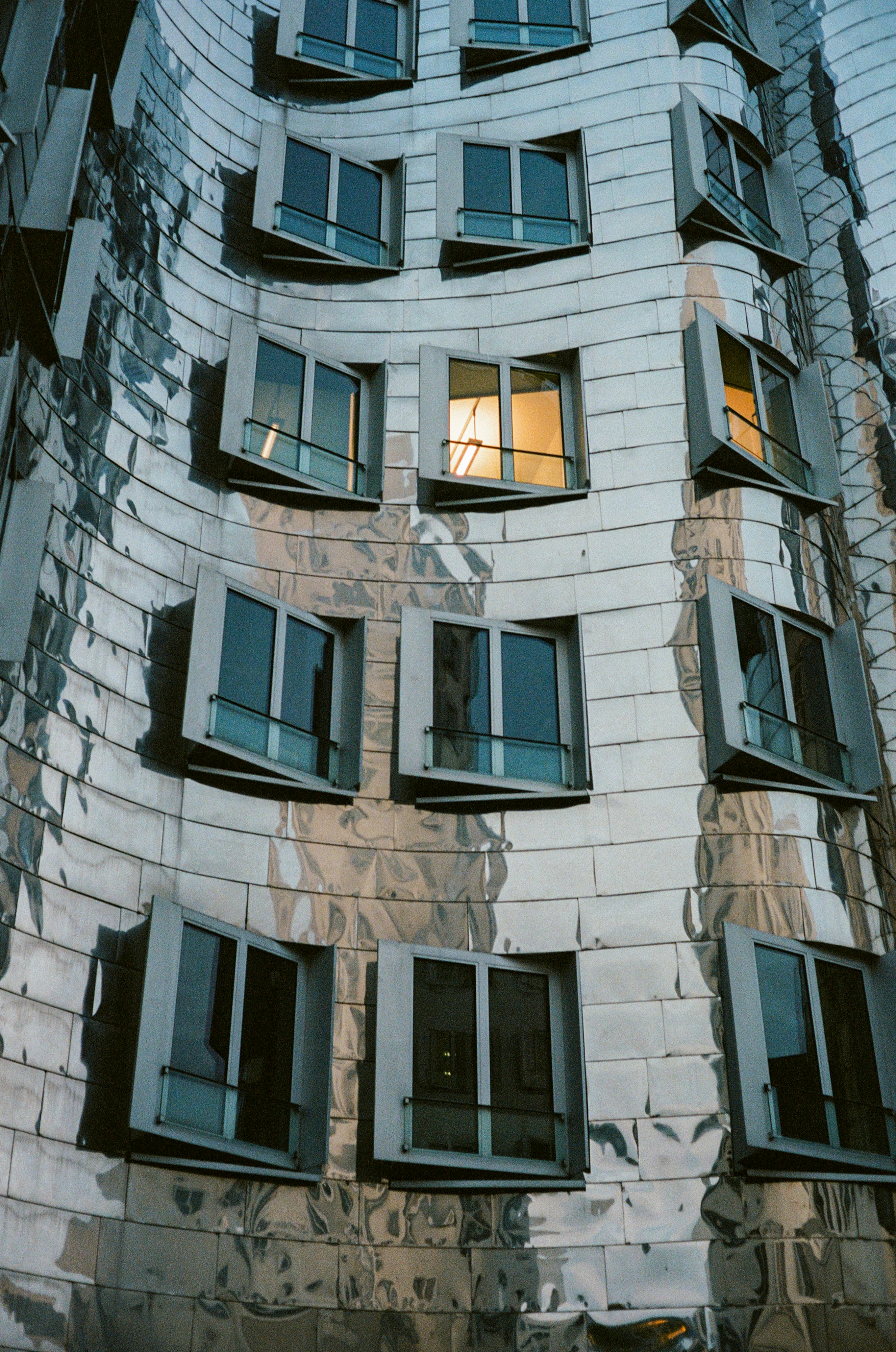 Reflective, curved metal panels form a modern facade of square windows. One interior window glows warmly, drawing the eye.