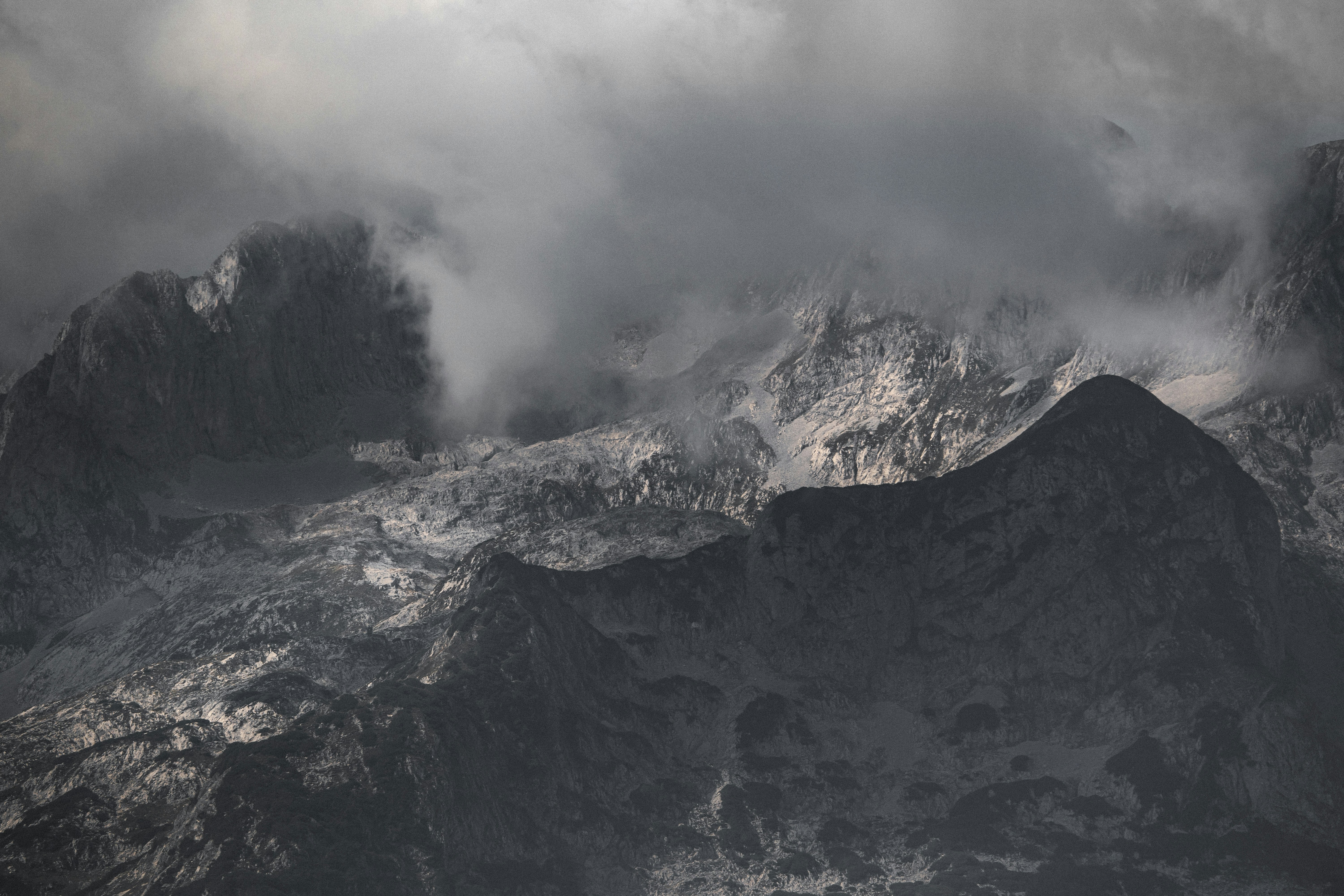 Mysterious mountain landscape shrouded in clouds, revealing rugged terrain and patches of snow. A dramatic interplay of light and shadow enhances the scene.