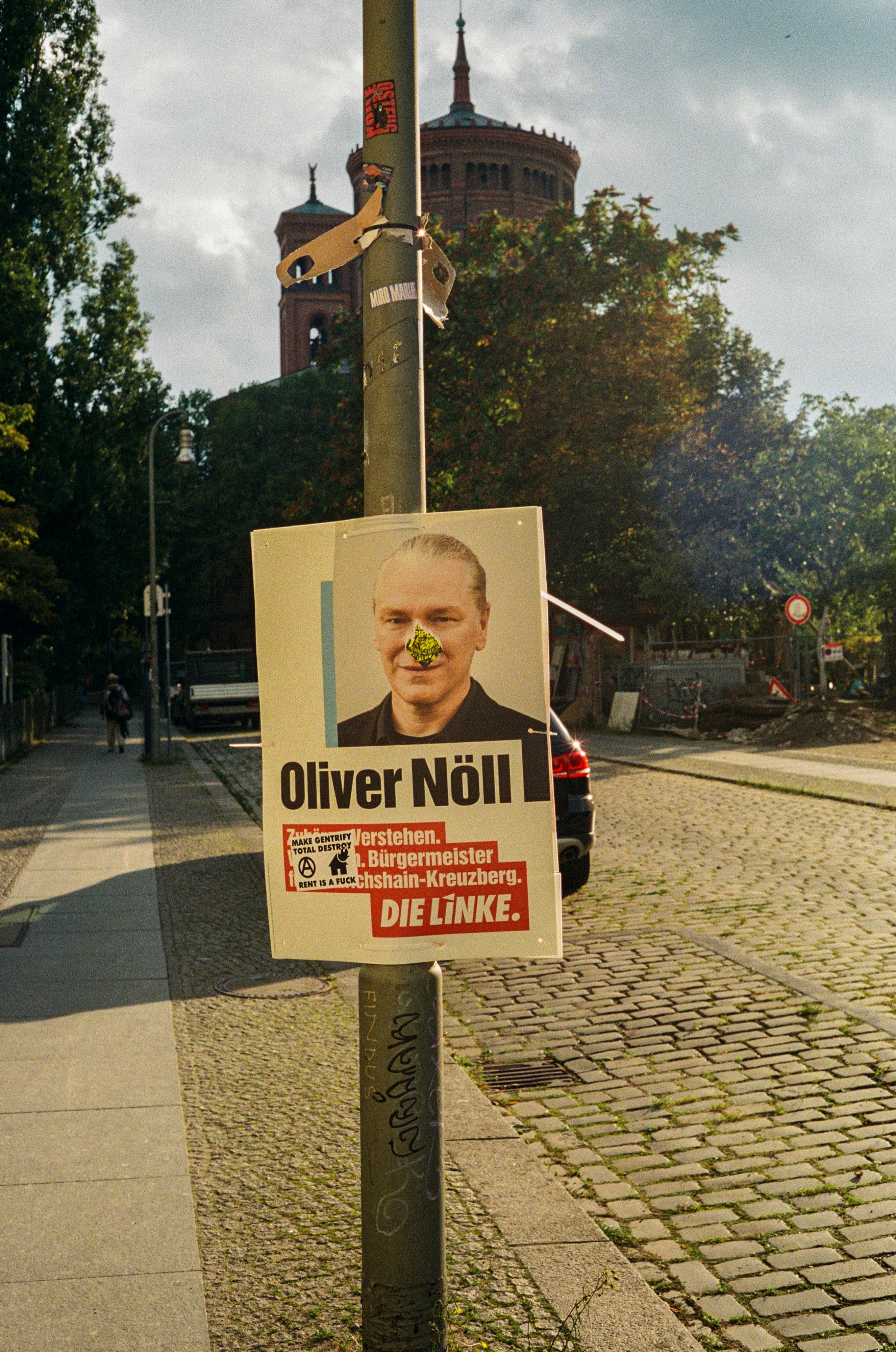 A political campaign poster for Oliver Nöll affixed to a weathered lamppost along a sunlit cobblestone street. The sign shows a portrait and Die Linke branding.