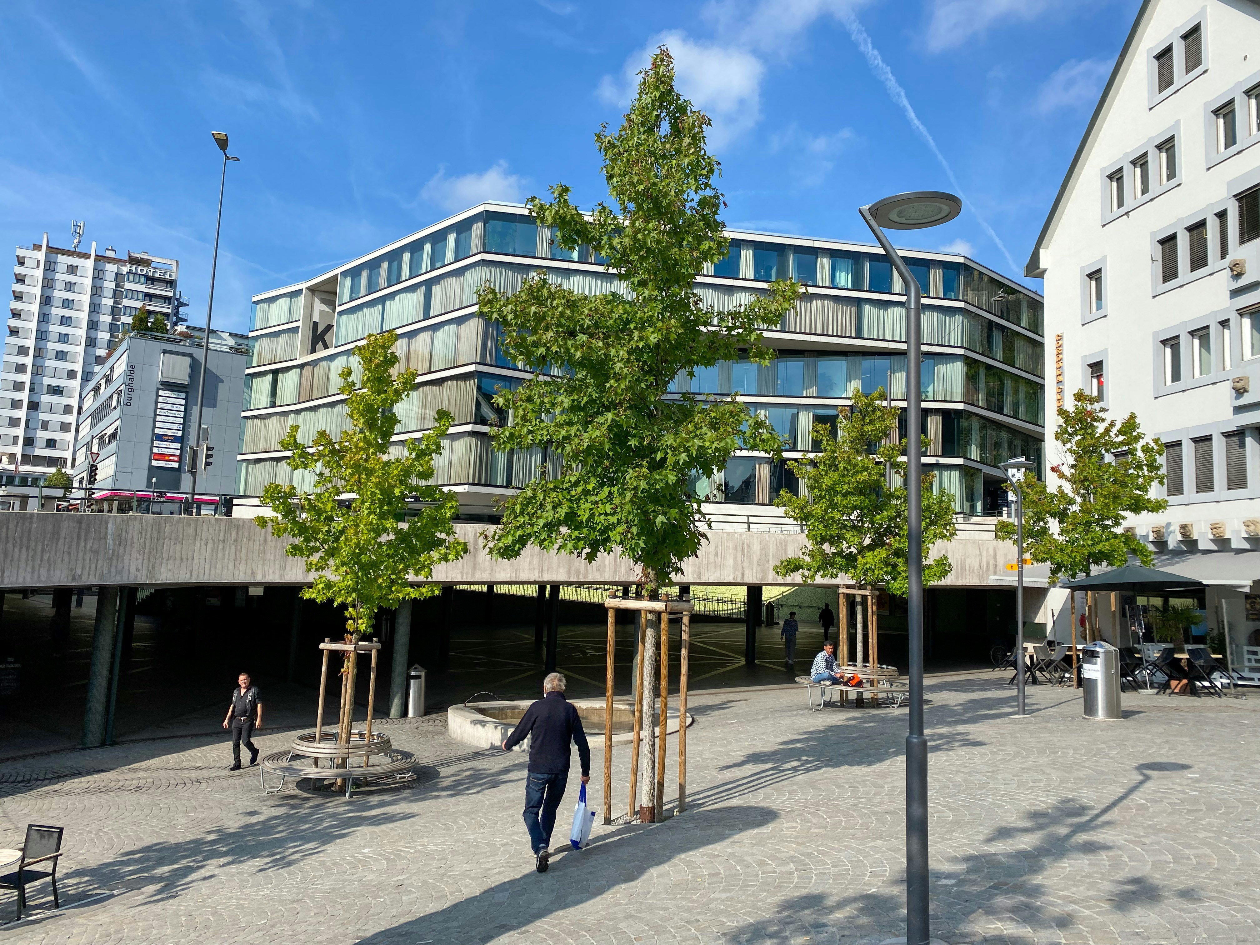 Man walking along a plaza bordered by modern architectural buildings under a clear blue sky.
