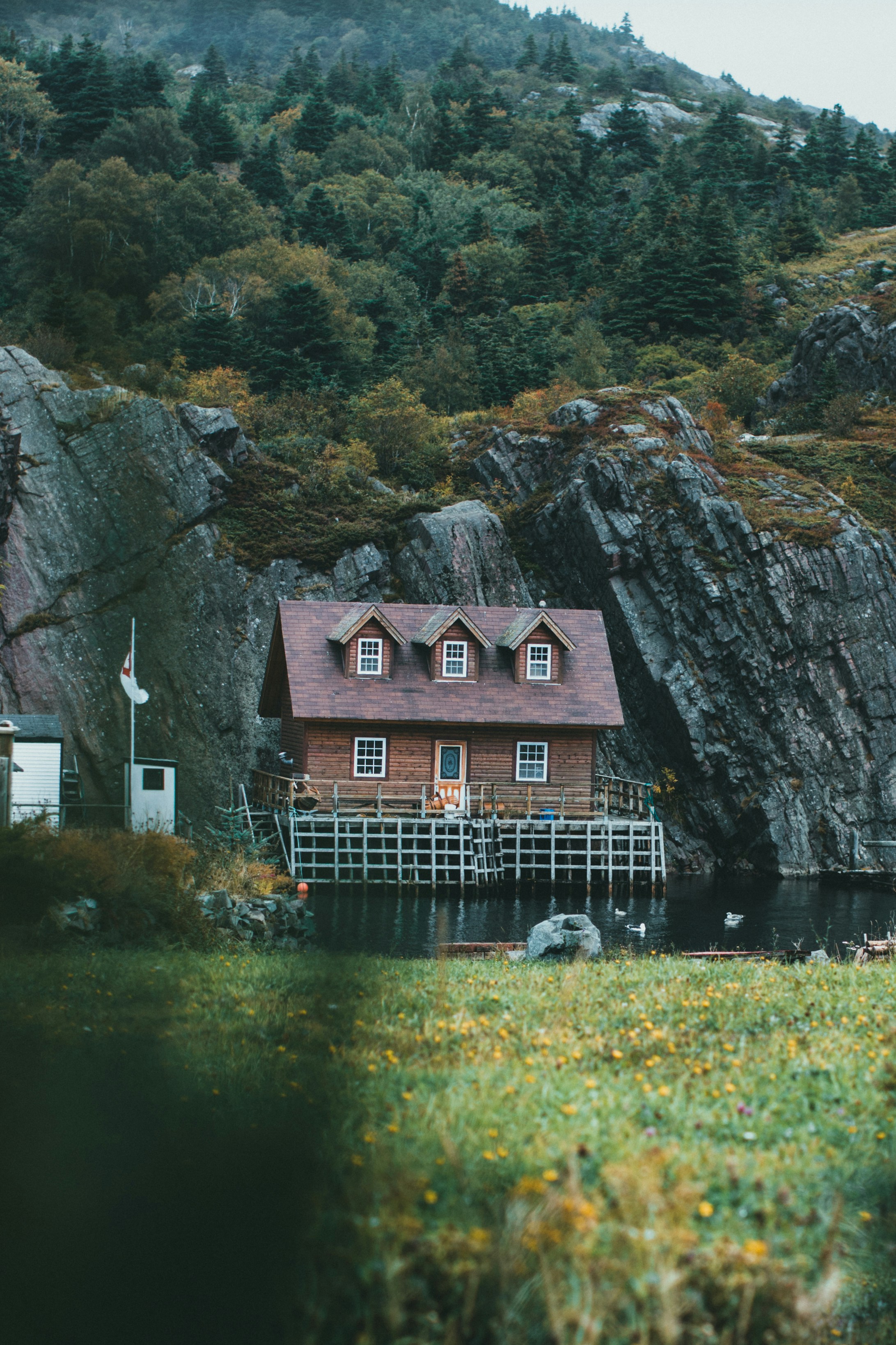 Charming wooden house perched on stilts beside a tranquil body of water, surrounded by autumn foliage and rocky cliffs.