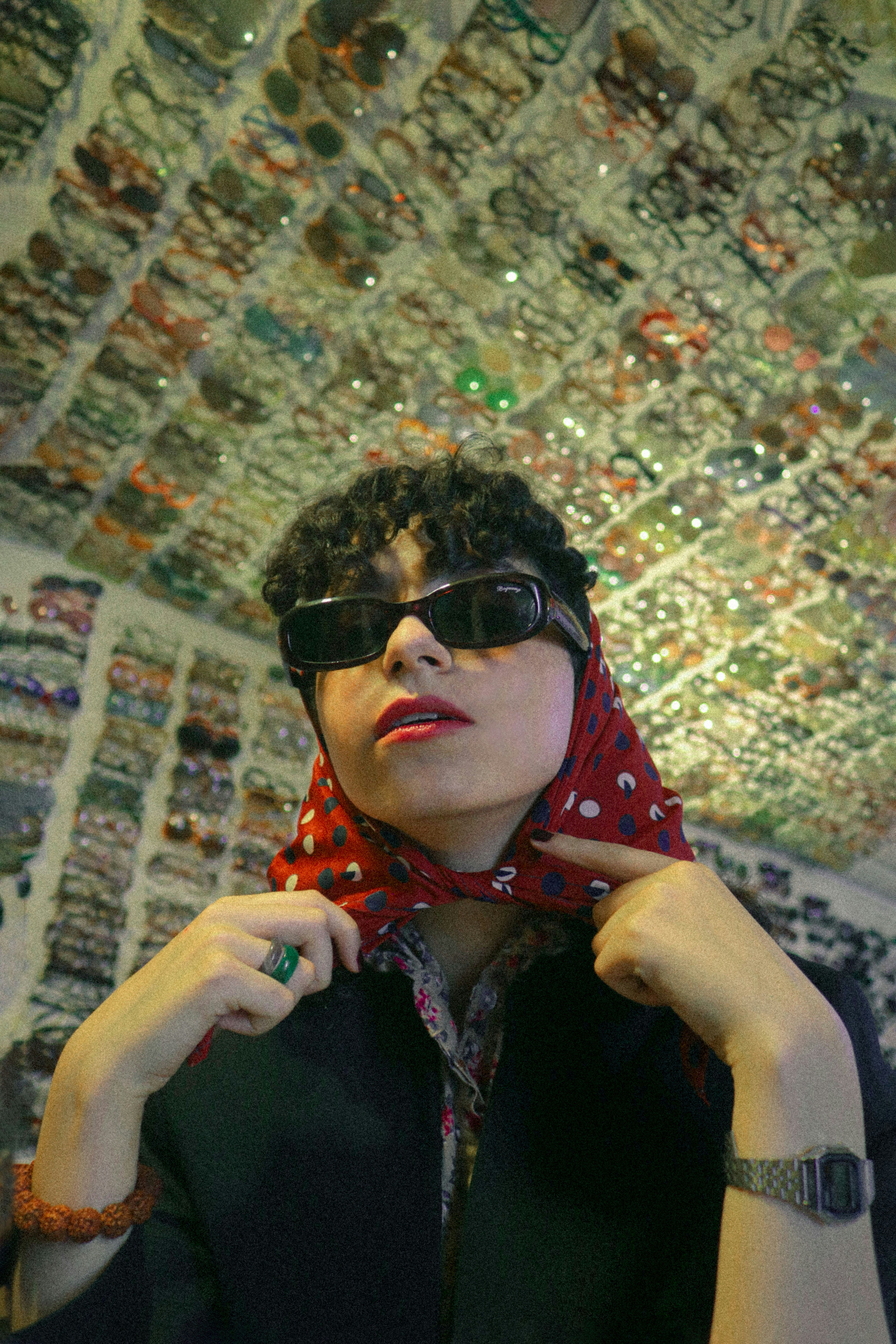 A fashionable individual wearing sunglasses and a polka-dotted scarf poses confidently in a room adorned with an array of eyeglasses. The eclectic backdrop adds a unique flair to the scene.