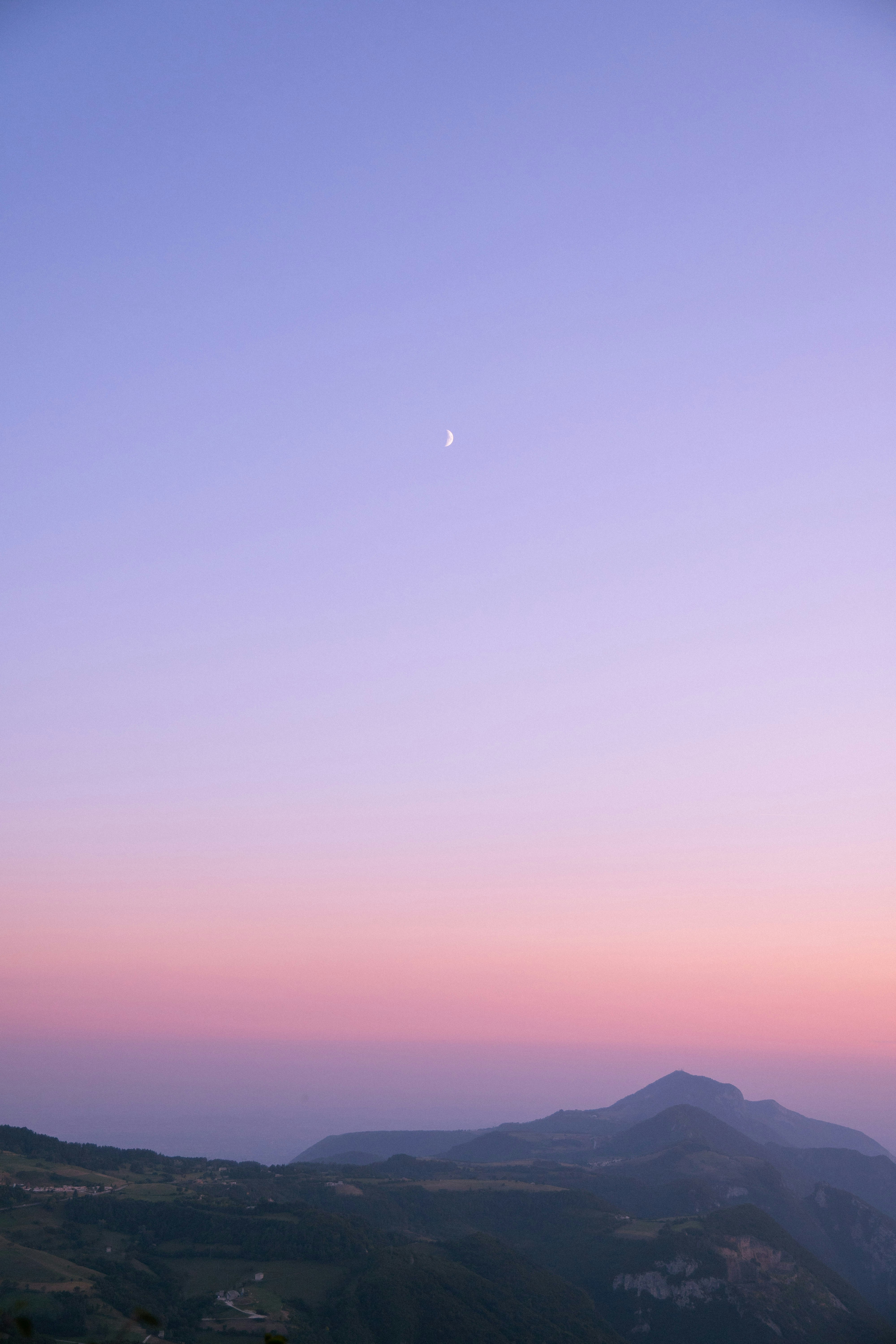 A crescent moon hangs delicately in a pastel sky above rolling hills at dusk, creating a serene atmosphere. The soft gradient of colors enhances the tranquil scene.