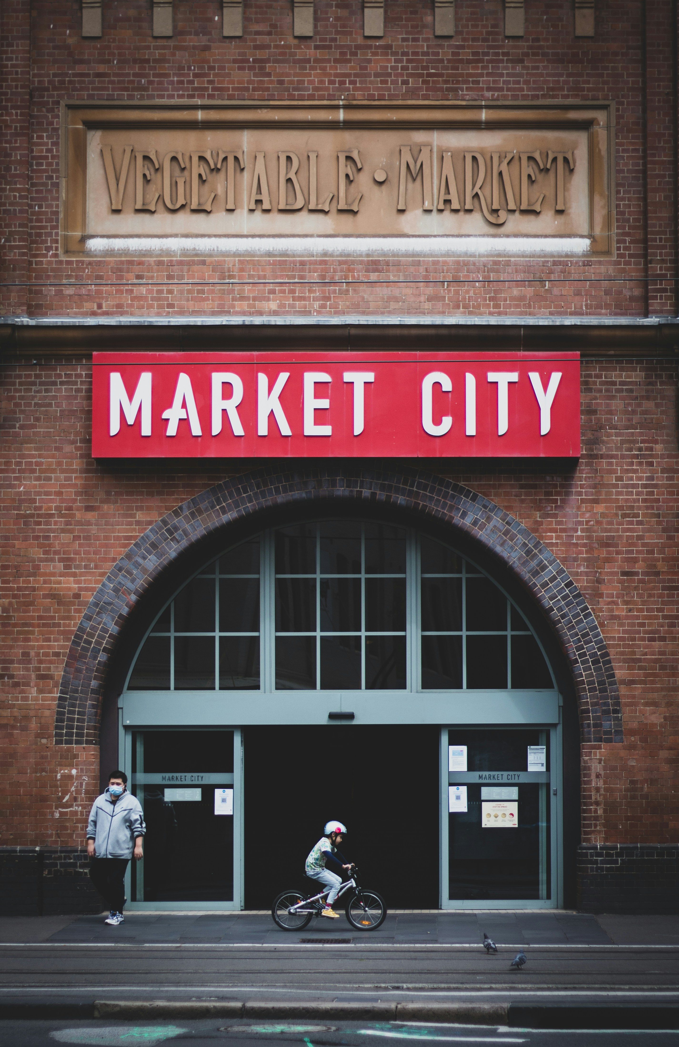 A cyclist rides a small bike past the brick Market City entrance beneath a bold red sign, while a masked pedestrian stands along the sidewalk.