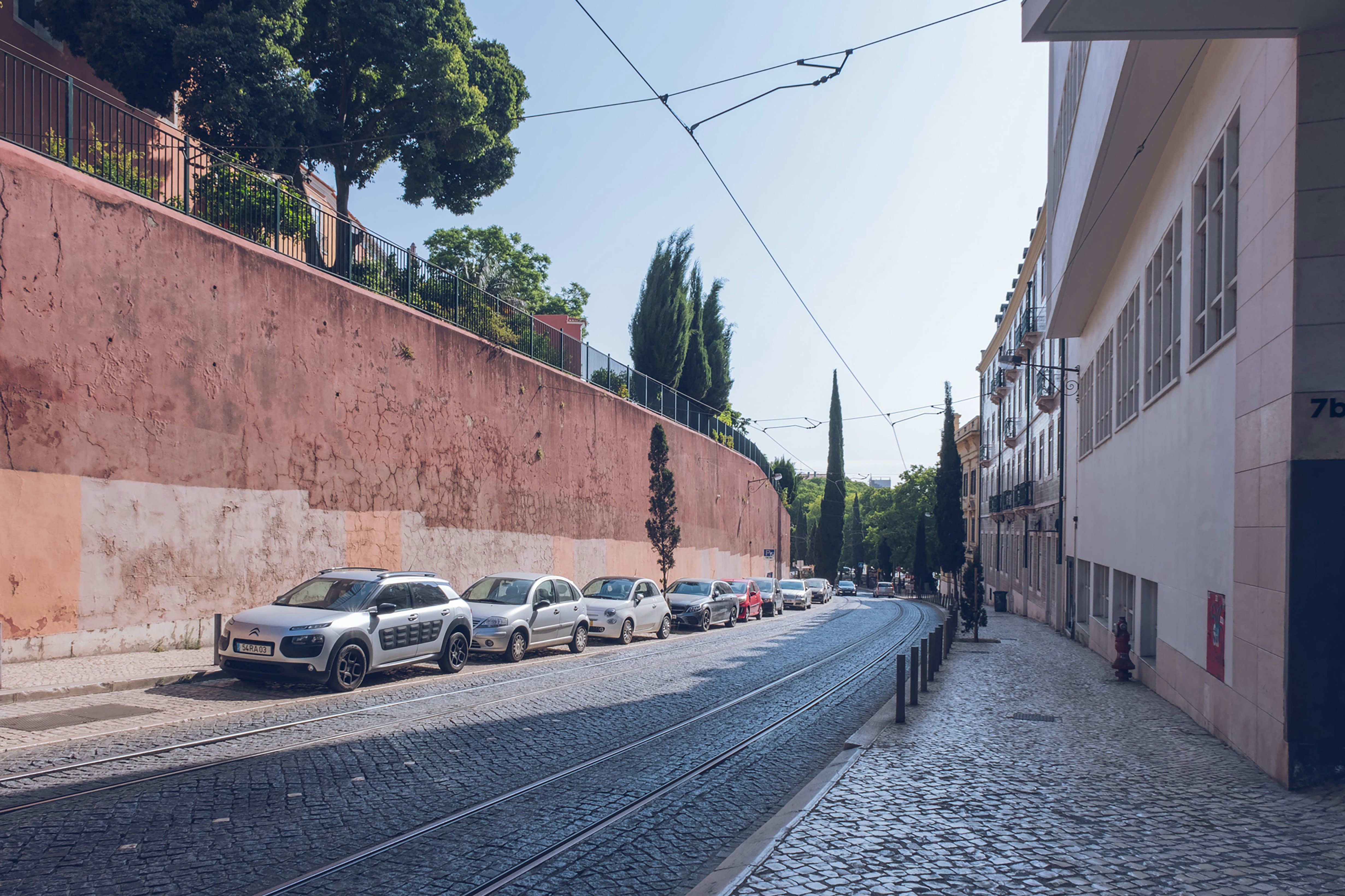 Row of cars lined up along a cobblestone street bordered by a red wall and lush trees.
