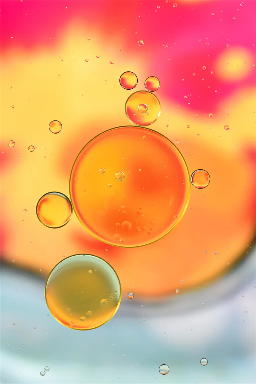 Translucent oil droplets float over a vivid pink, orange, and yellow background with a few smaller bubbles scattered around the frame. The largest droplet sits near the center as the main visual anchor.