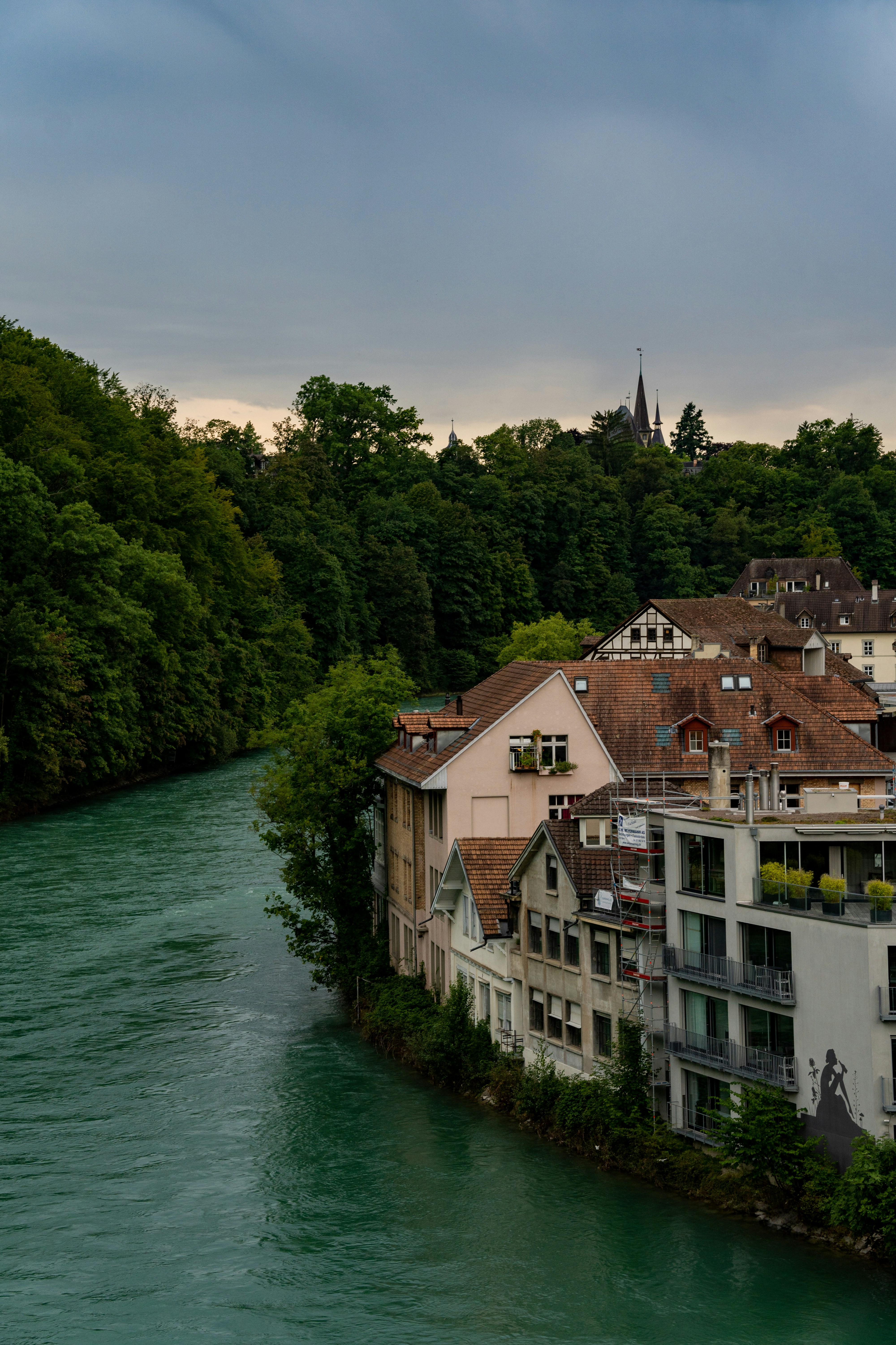 Charming riverside architecture nestled among lush greenery, with a distant church steeple peeking through the trees. The tranquil river flows gracefully alongside the buildings.