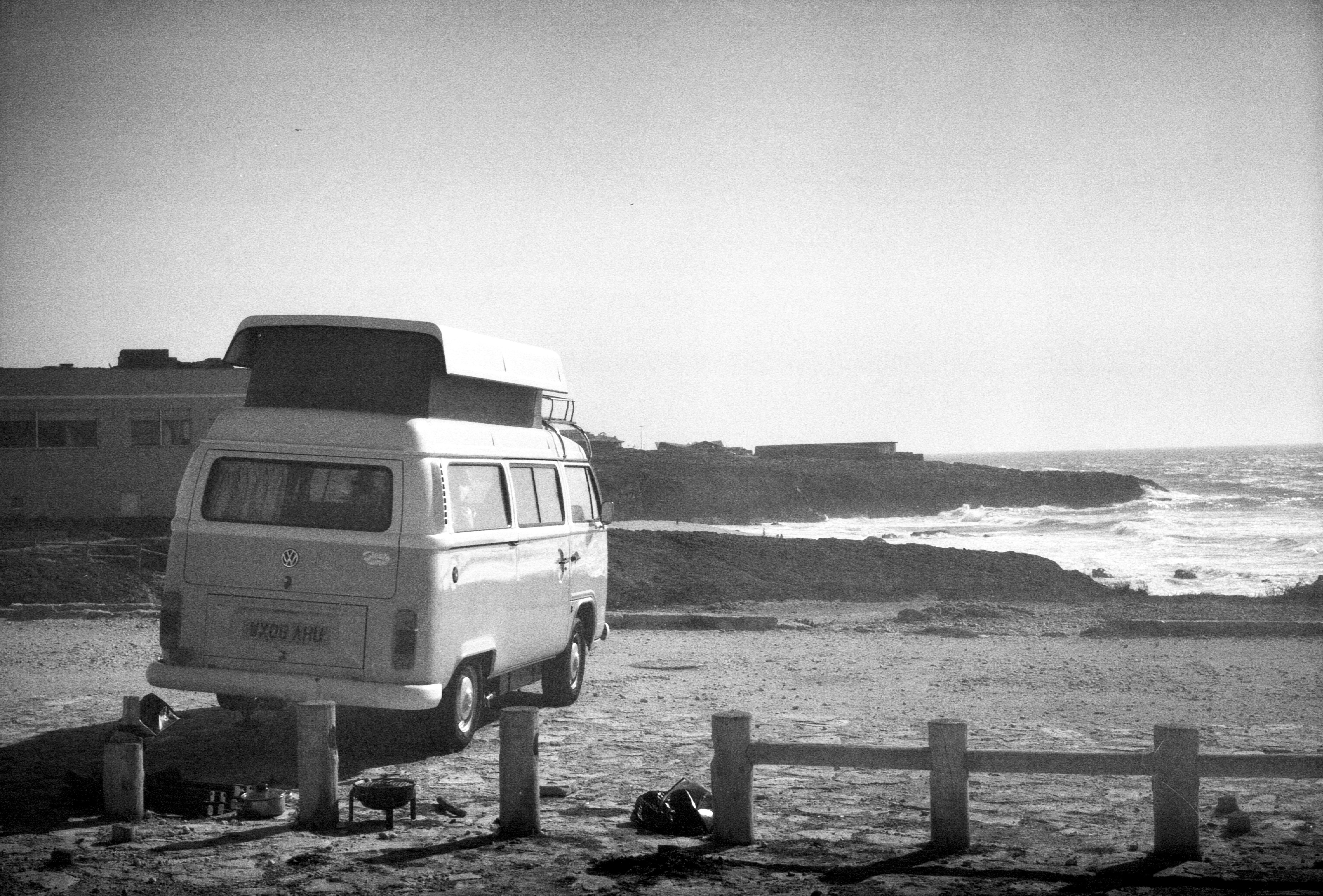 Monochrome photograph of a vintage VW camper van parked on a sandy beach with a weathered fence nearby. Distant waves roll along a rocky shoreline in the background.