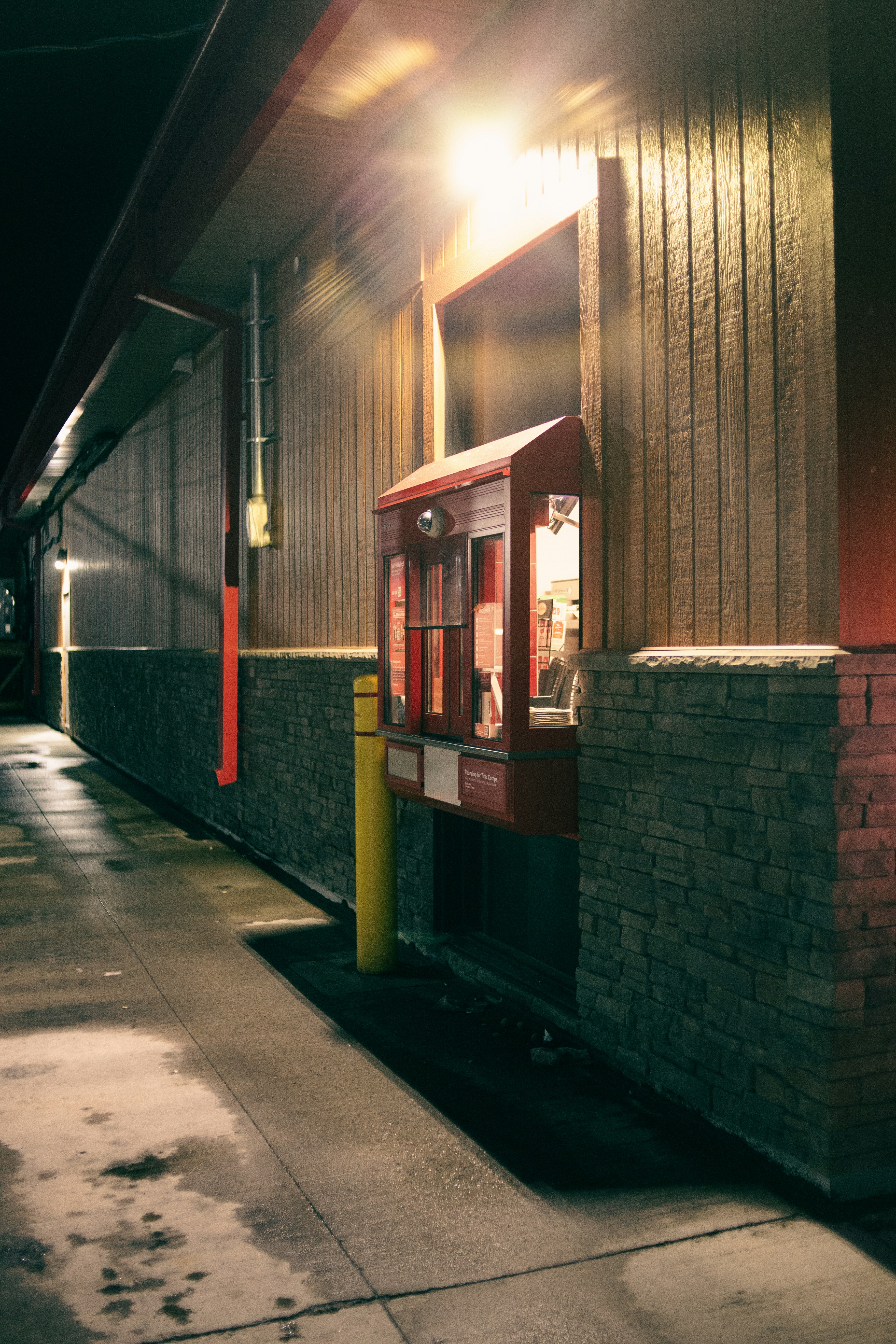 A brightly lit drive-thru window at night, showcasing a vibrant red ordering booth against a textured wall. The wet pavement reflects the ambient light.