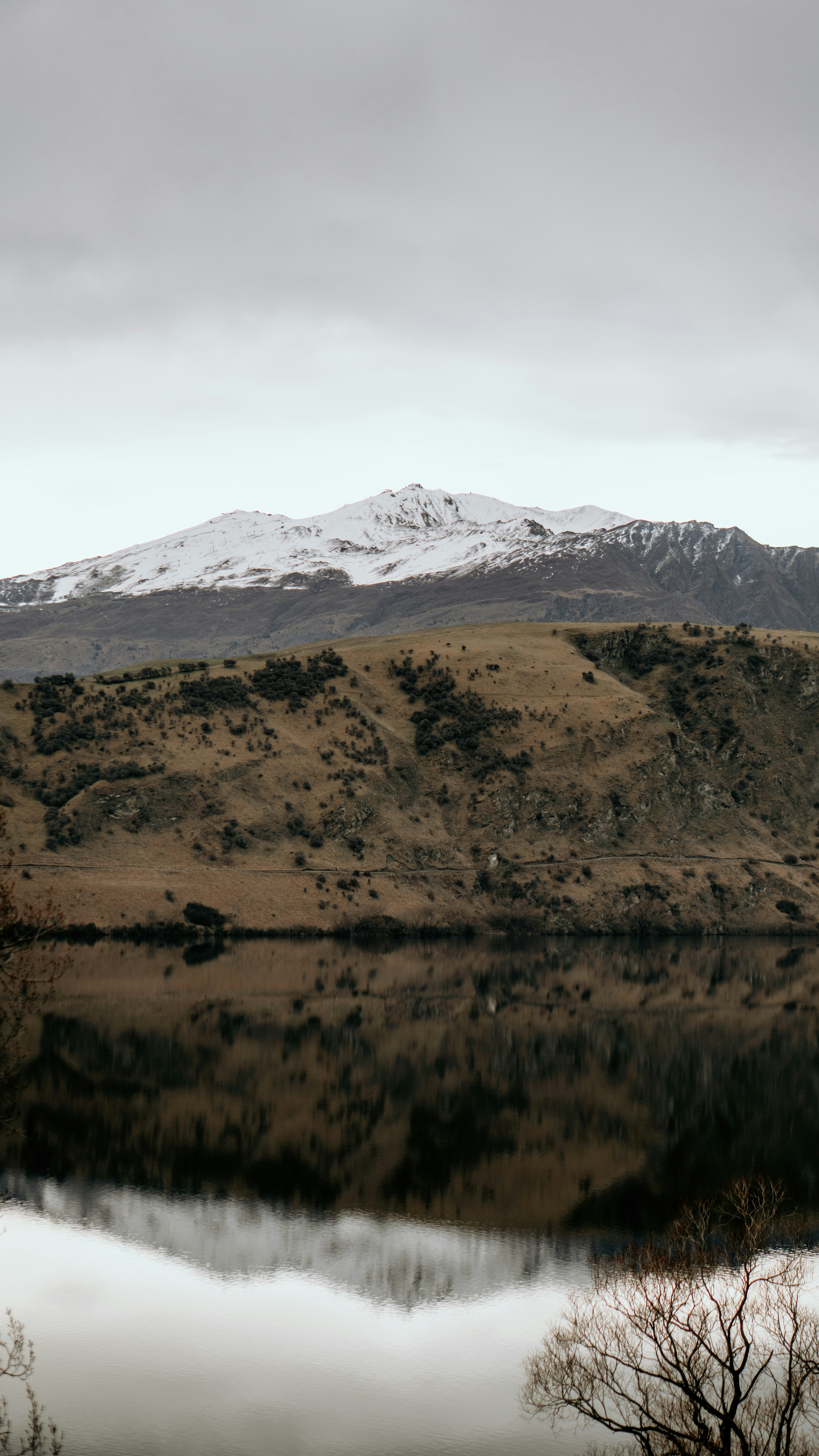 Snow-capped mountains mirrored in a tranquil lake, surrounded by rugged terrain and soft clouds. The scene captures the essence of nature's calm beauty.
