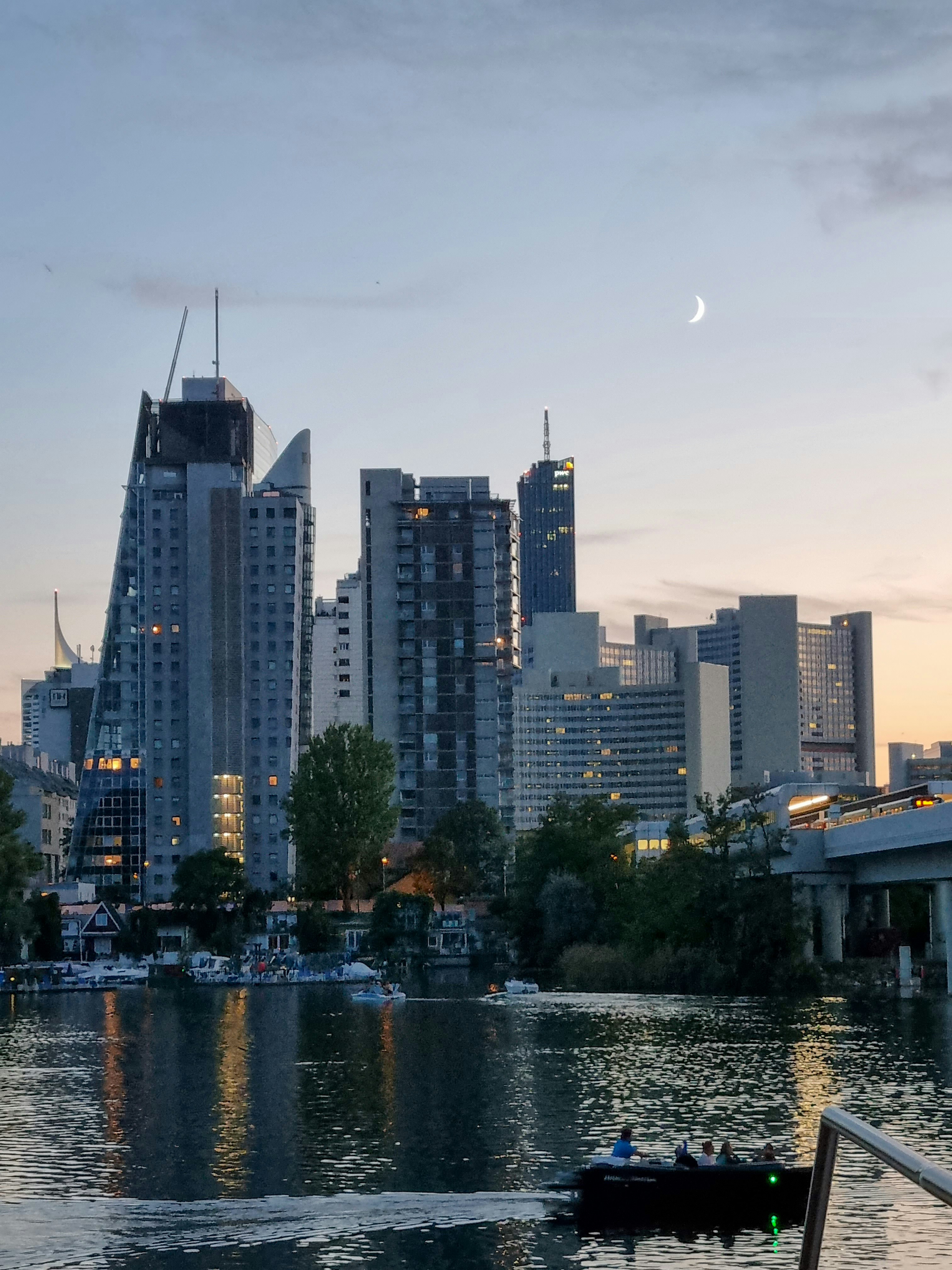 Cityscape photograph of a glass-clad skyline along a calm river at dusk, with a crescent moon in the sky.