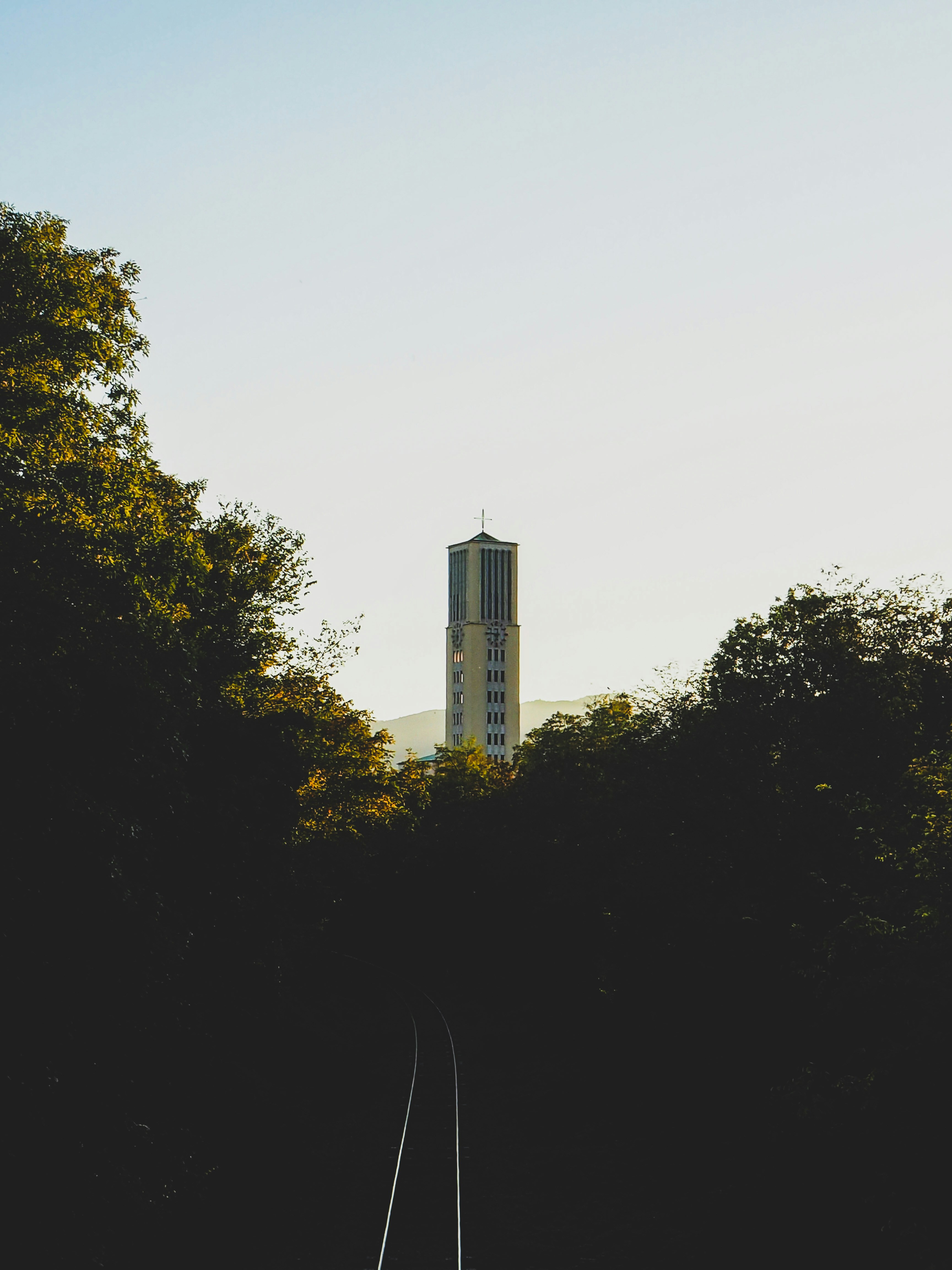 A tall clock tower emerges from a green landscape, framed by dense foliage and a clear sky. The scene captures a serene moment along a railway track.