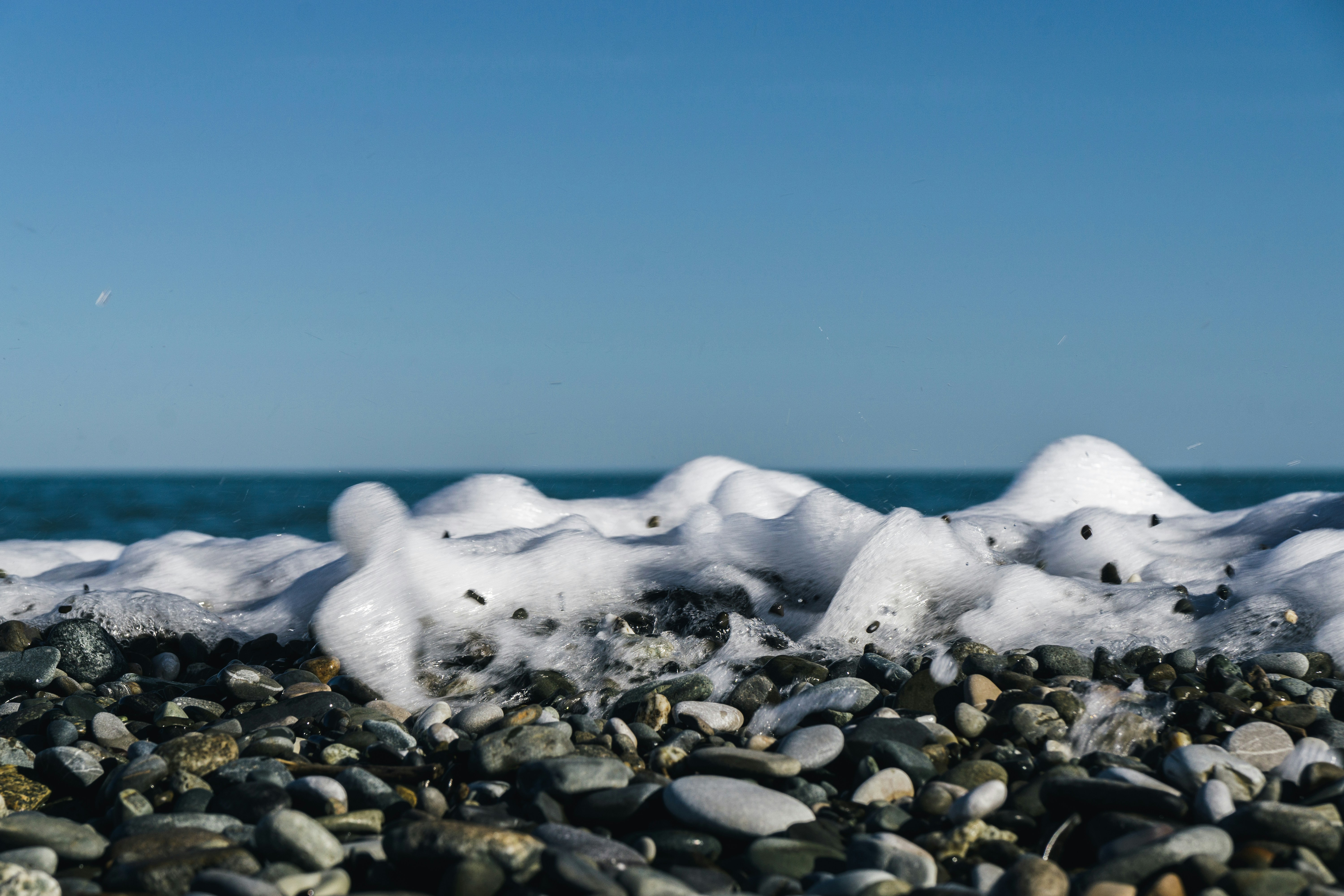 Foam-Crowned Pebbles: A Quiet Shoreline Under Arctic-Blue Skies