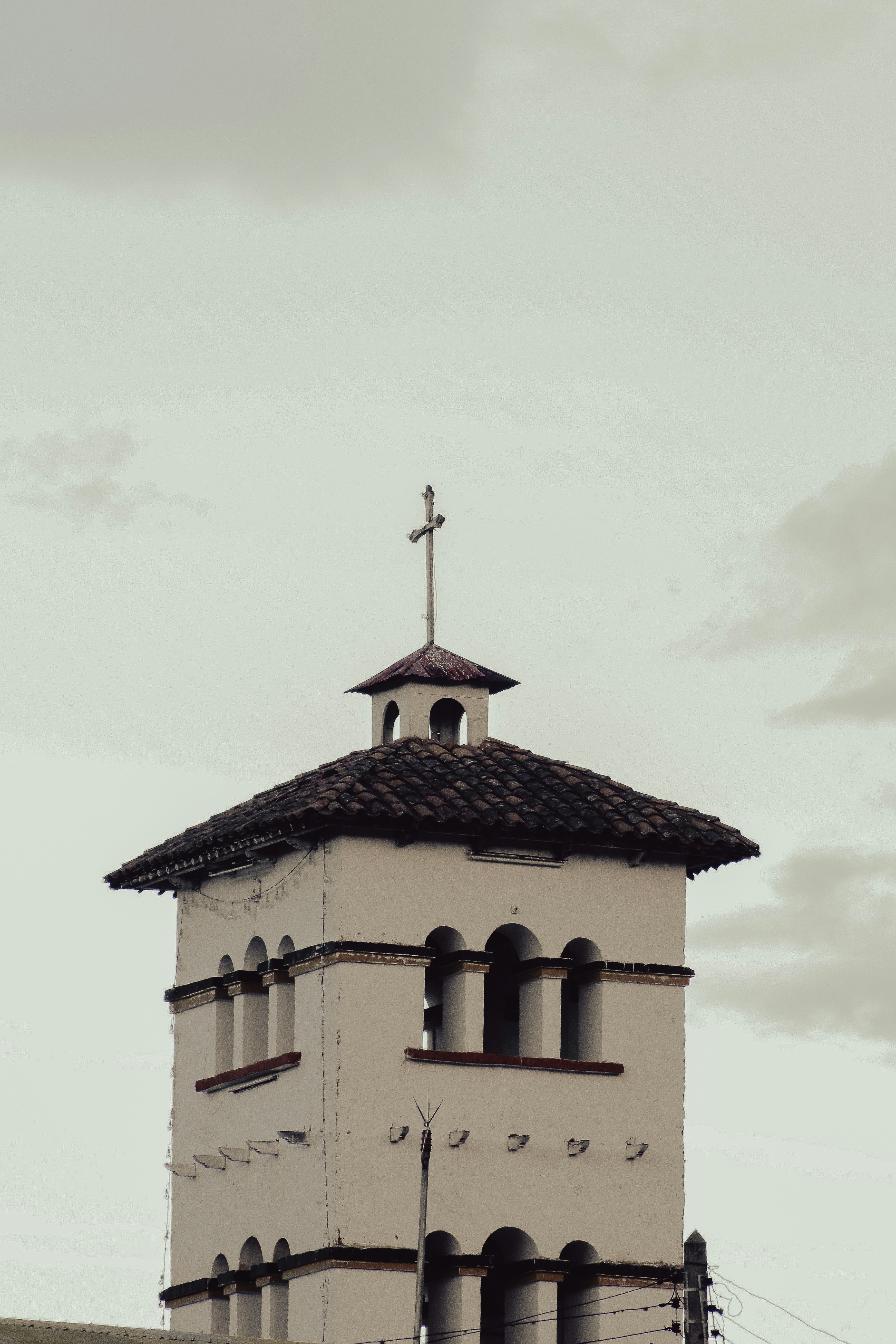 Historic bell tower with arched windows and a cross-topped roof under a cloudy sky.