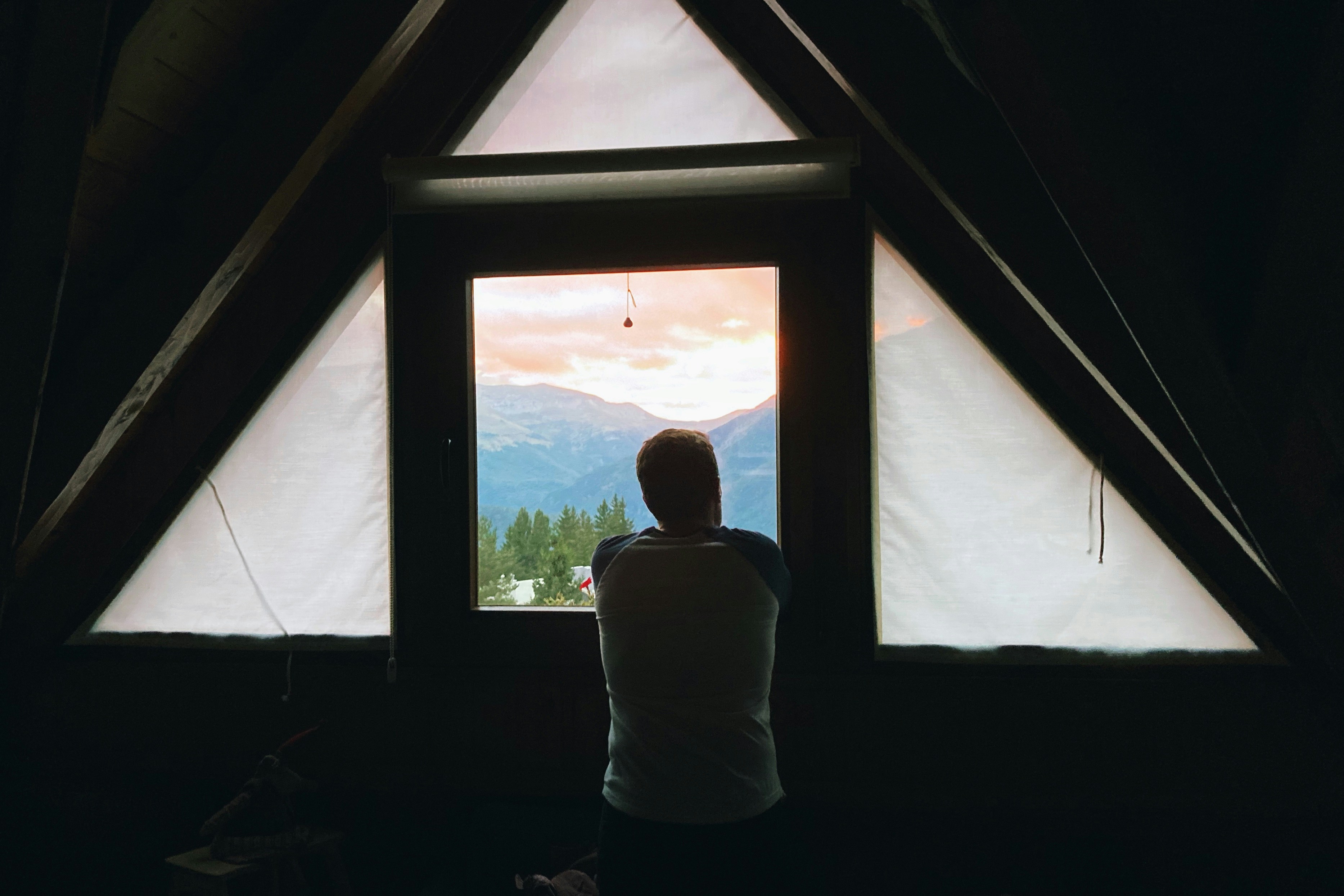 Person gazing out of a triangular window, framed by wooden beams, as the sun sets over a mountainous landscape.