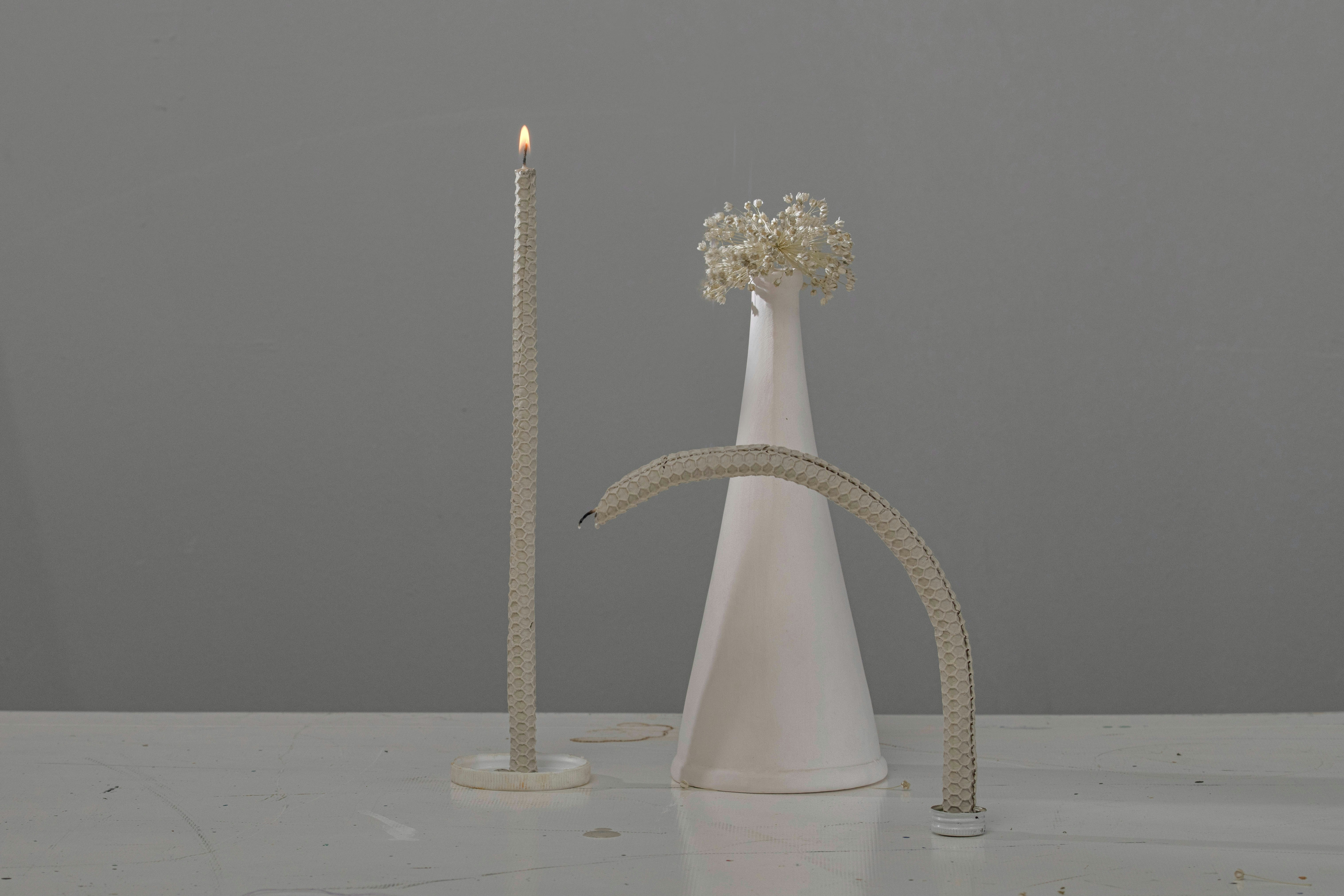 A minimalist arrangement featuring a lit candle, a curved decorative element, and a delicate floral vase against a soft gray background.