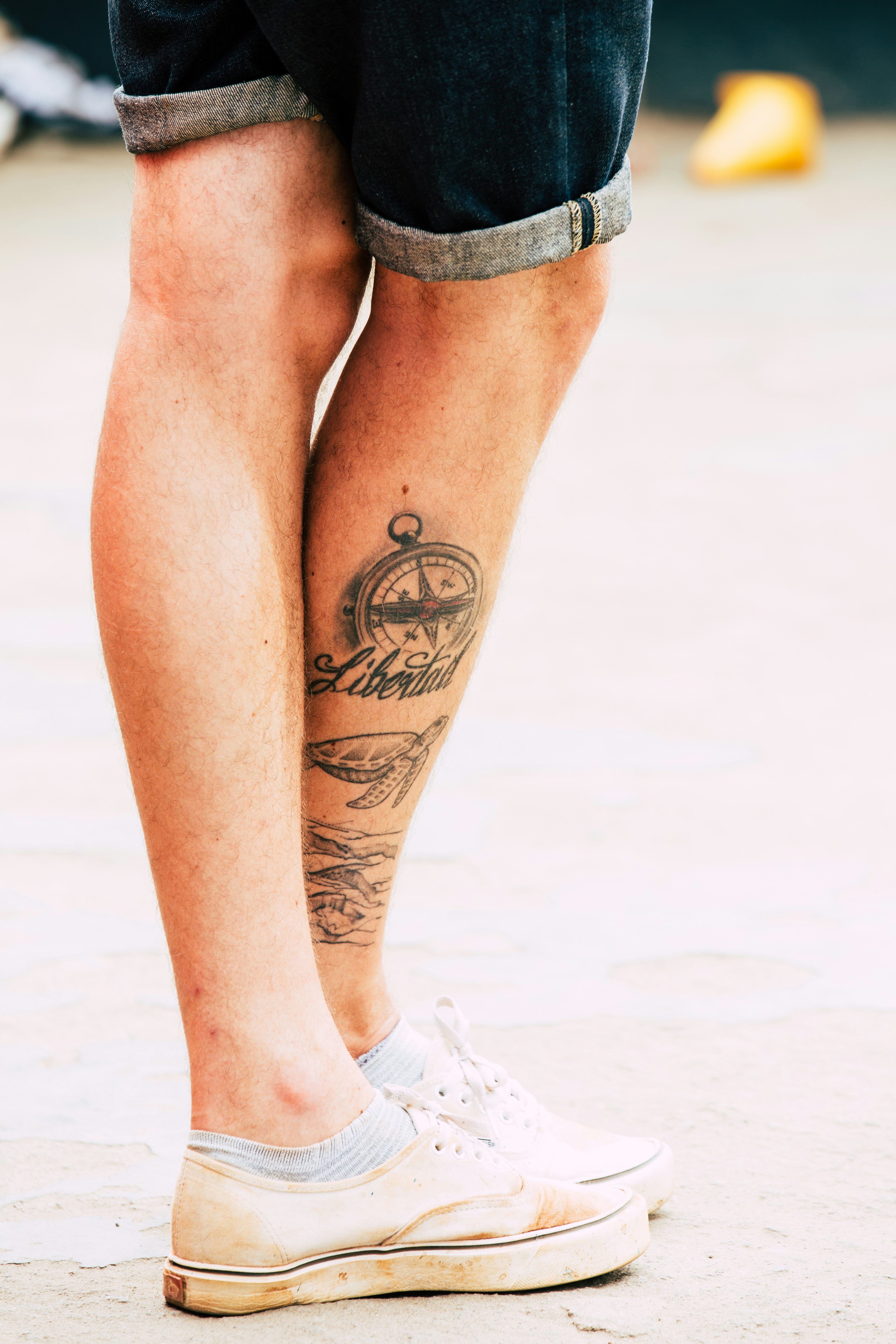 Close-up of a leg featuring a detailed compass tattoo, paired with worn white sneakers and rolled denim shorts.