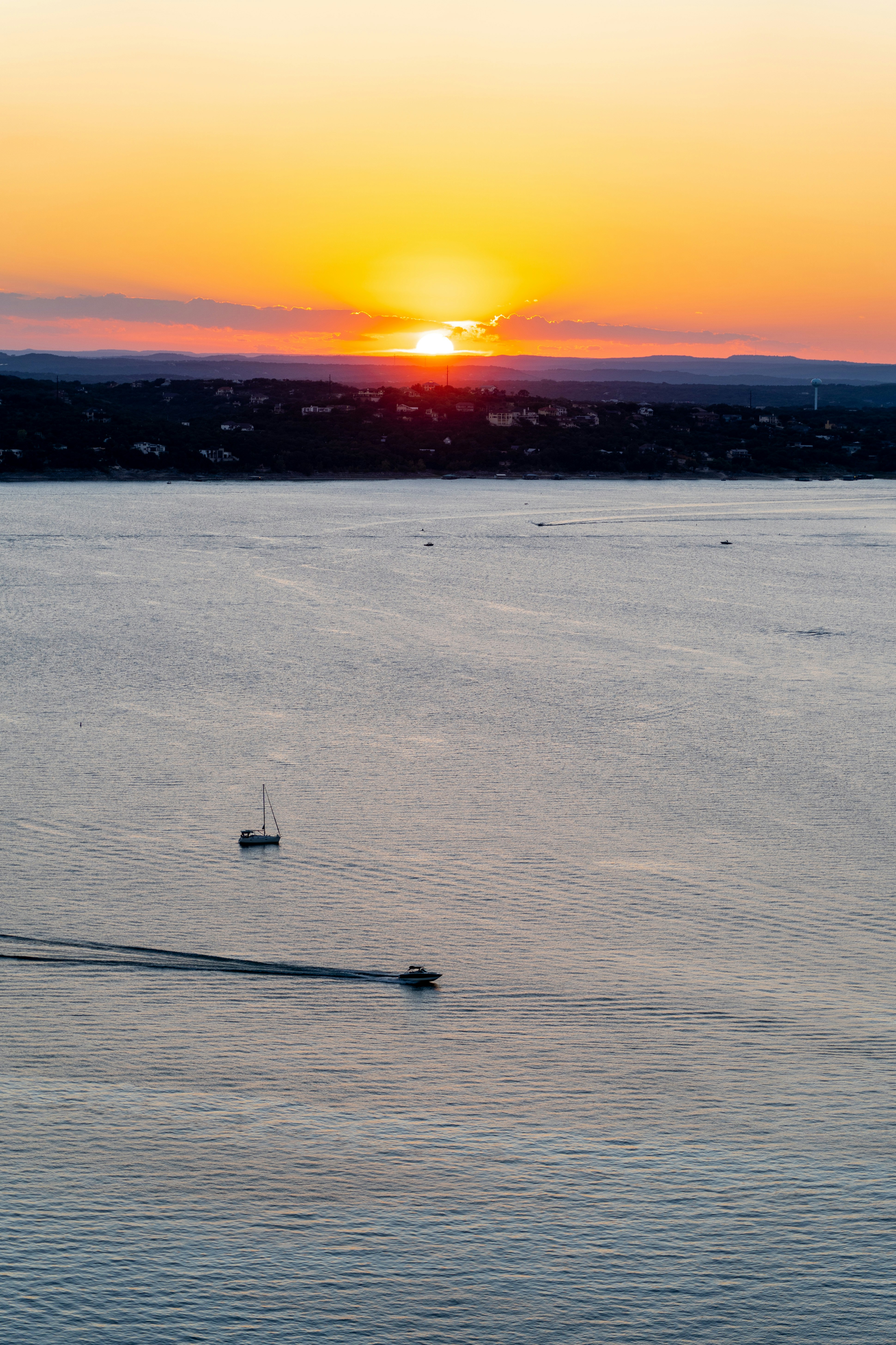 Boats glide across Lake Travis as the sun sets behind distant hills, casting an orange glow on the water.