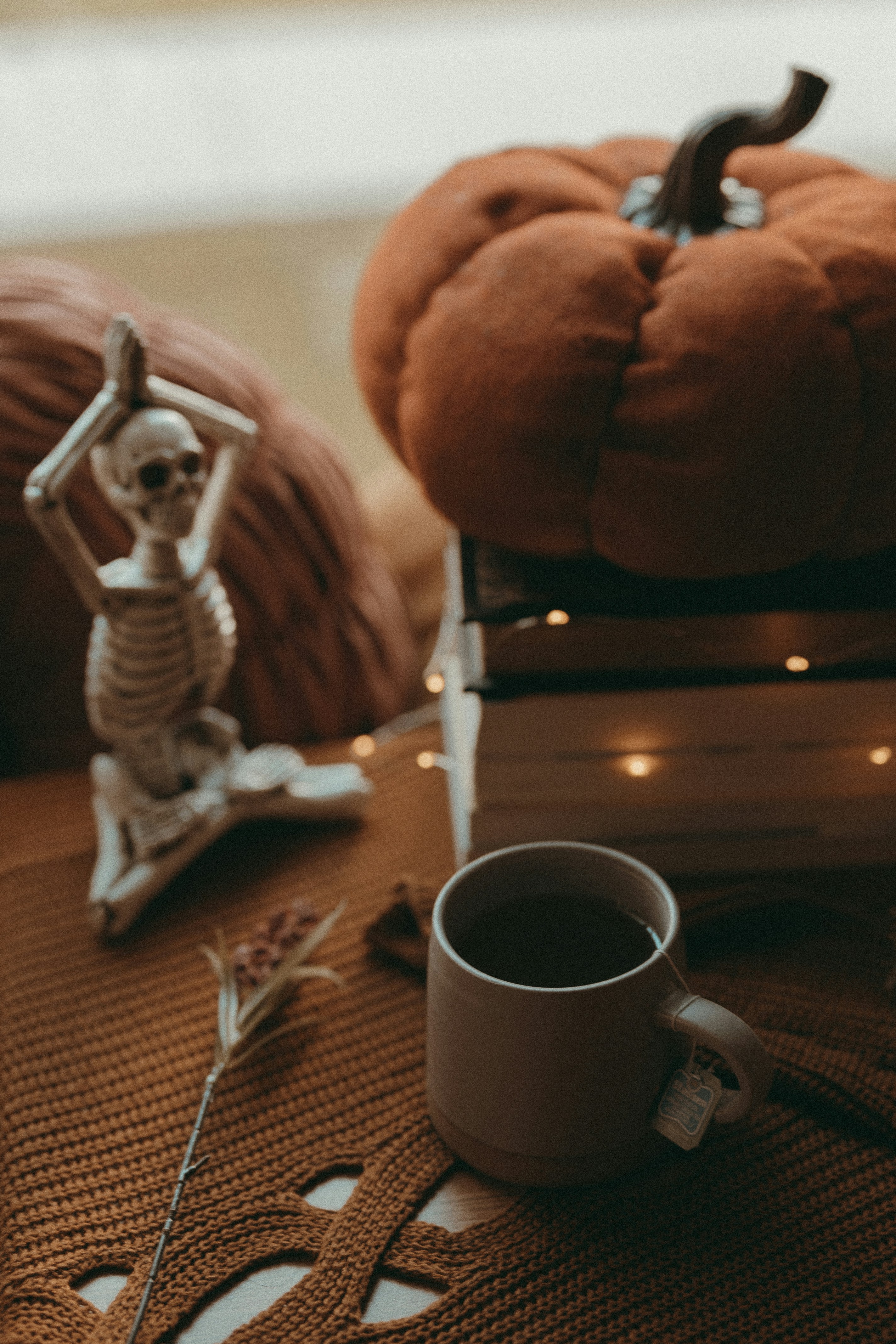 A whimsical skeleton in a yoga pose sits beside a warm mug of coffee and a plush pumpkin, set against a textured tablecloth with autumnal hues.