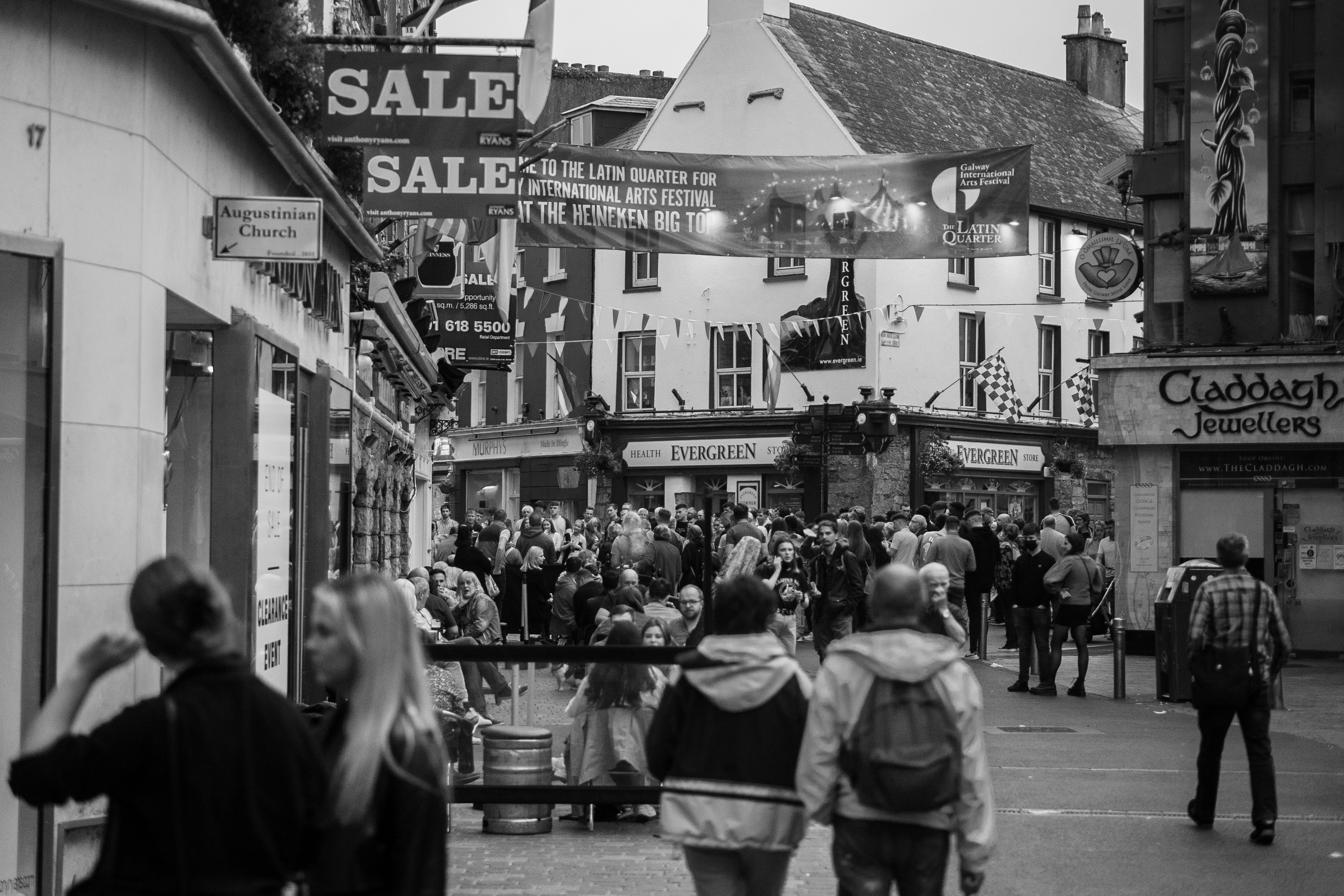 Crowded street scene with various sale signs in a lively shopping district, captured in black and white.