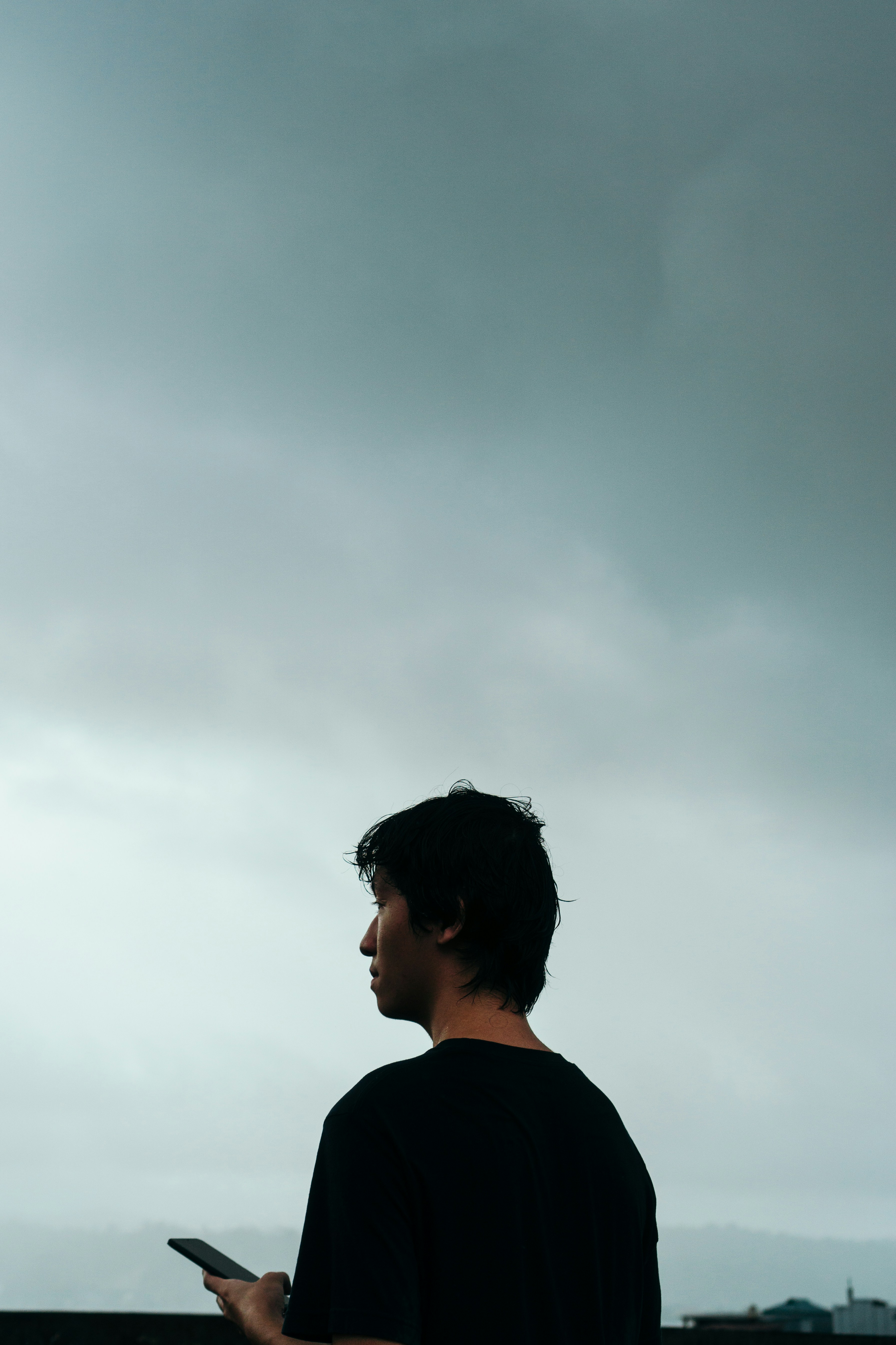 Silhouette of a young man standing against a moody sky, holding a smartphone, embodying a moment of reflection.