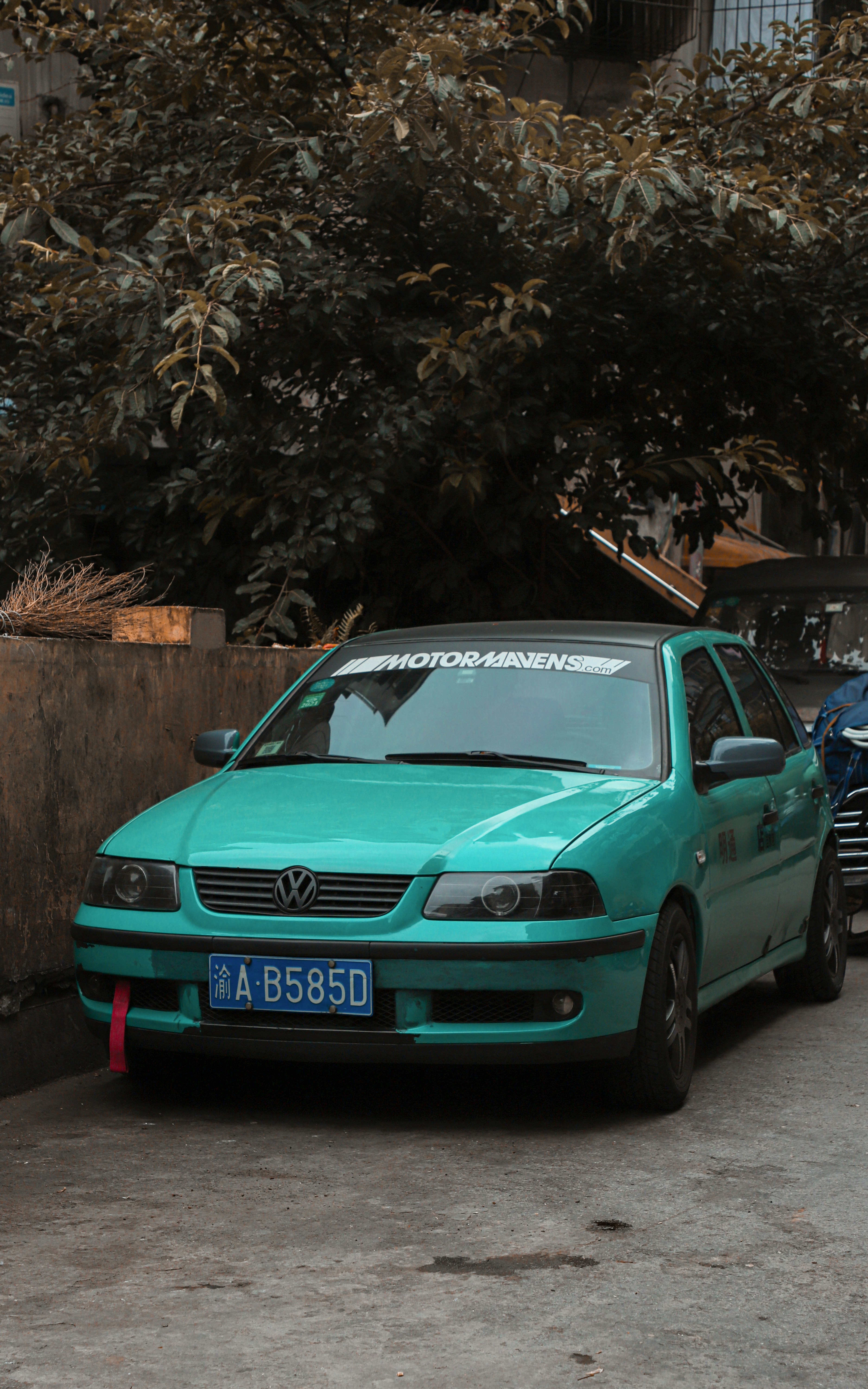 A vibrant aqua Volkswagen parked in a narrow alley, surrounded by lush foliage and urban elements.