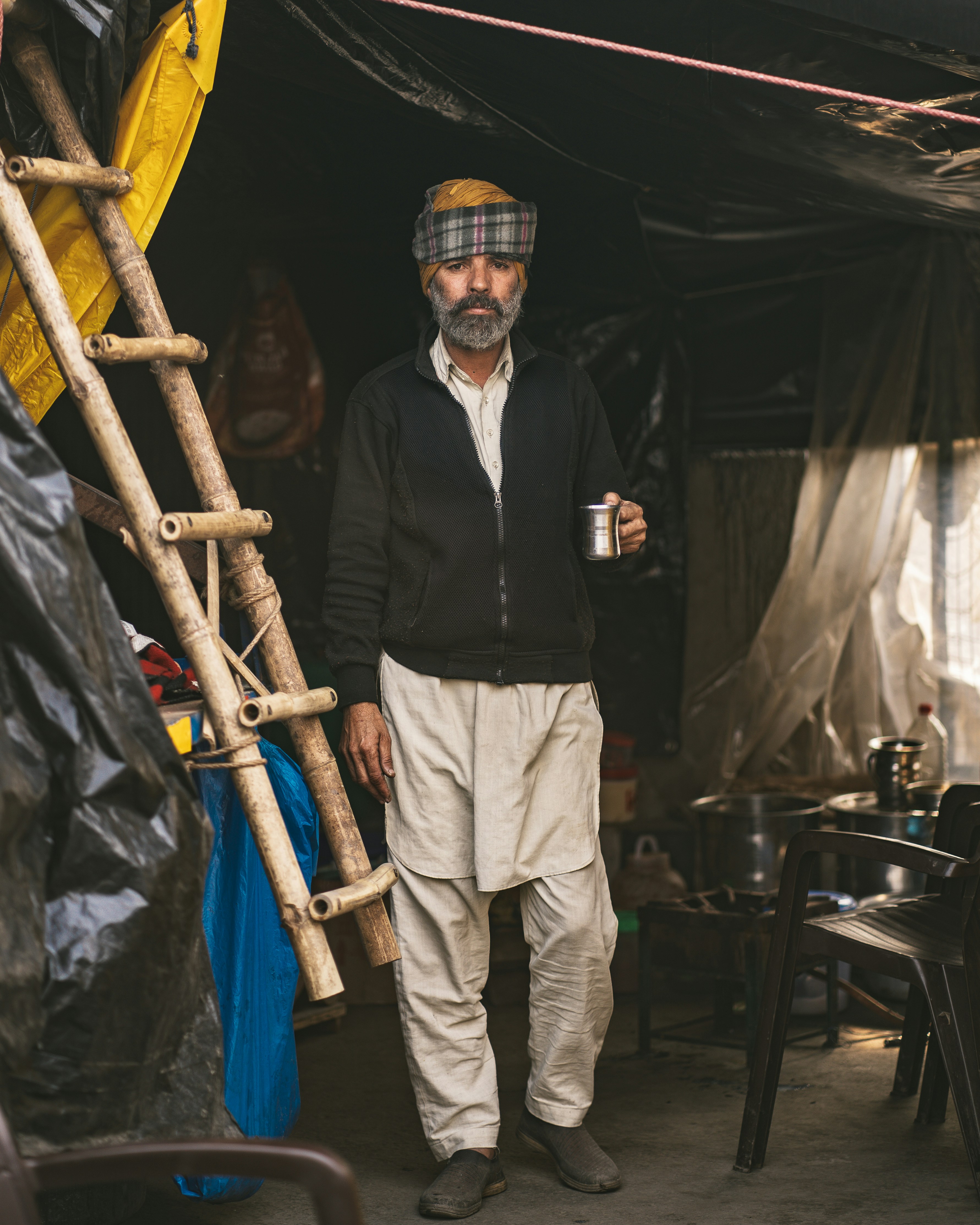 An elderly man holding a cup stands in a rustic setting, surrounded by simple furnishings and a ladder, embodying a sense of warmth and tradition.
