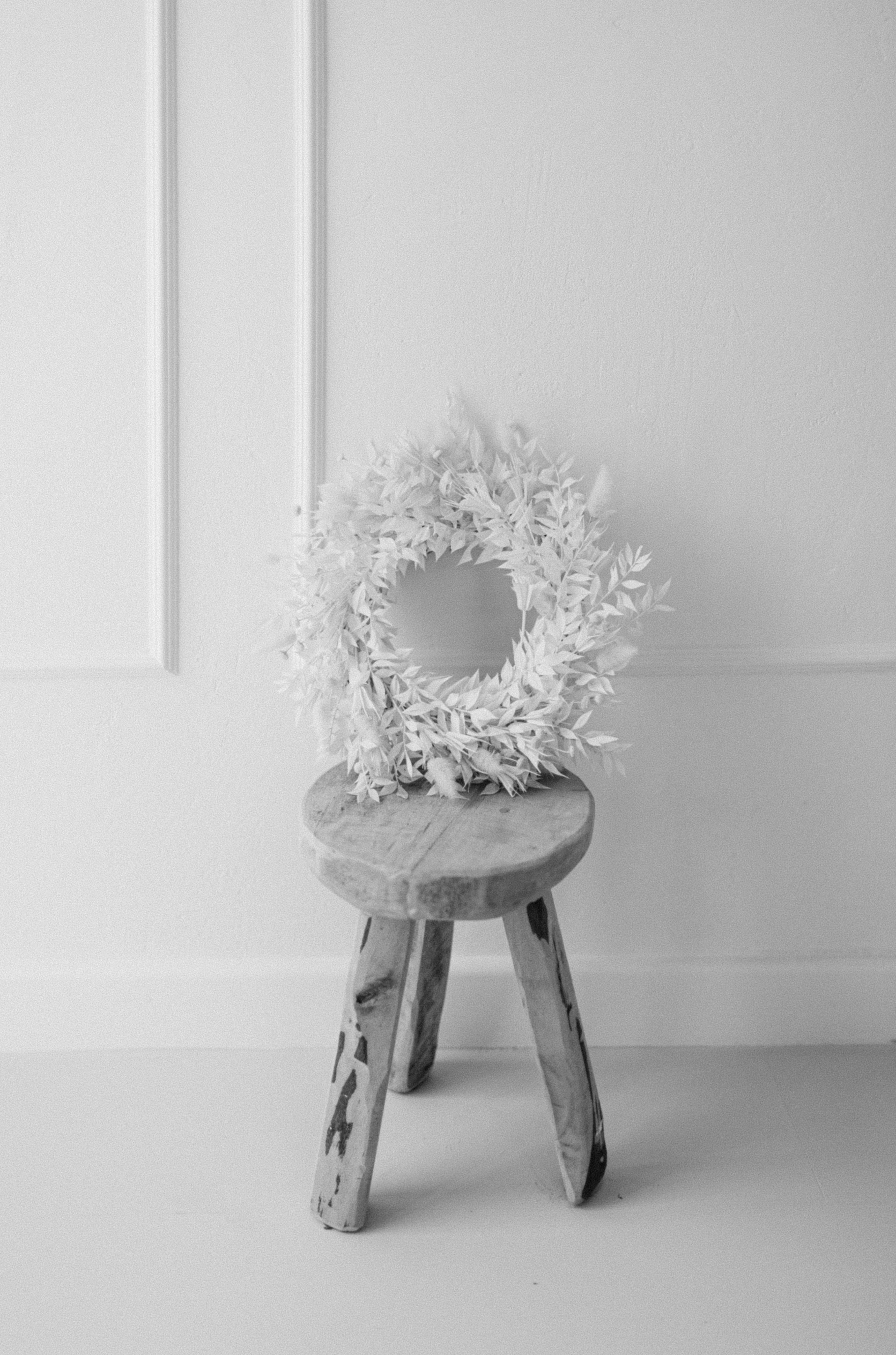 Delicate white wreath resting on a rustic wooden stool against a soft, neutral backdrop.