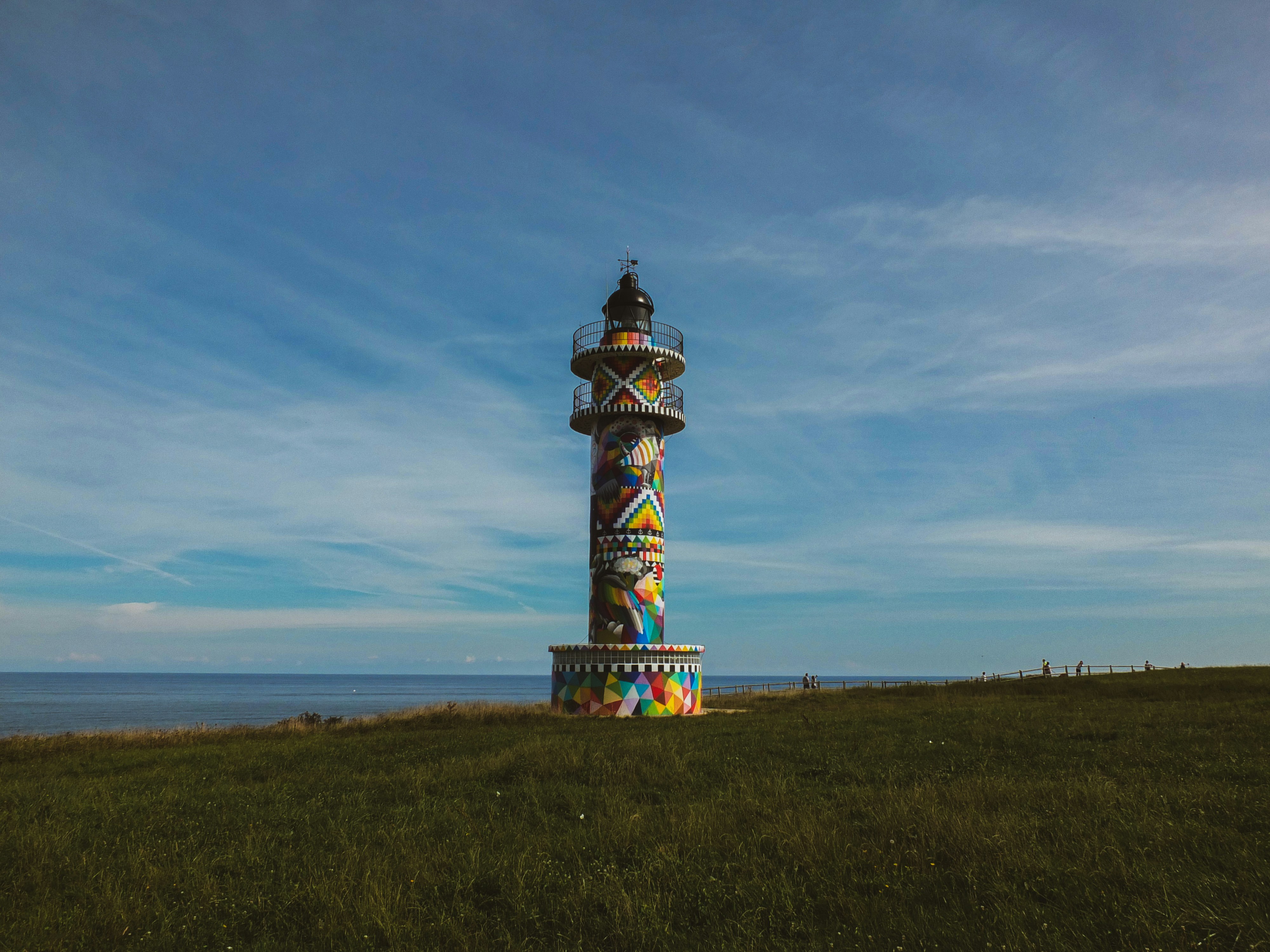 Colorful lighthouse rises from a grassy bluff with the sea and blue sky in the background.